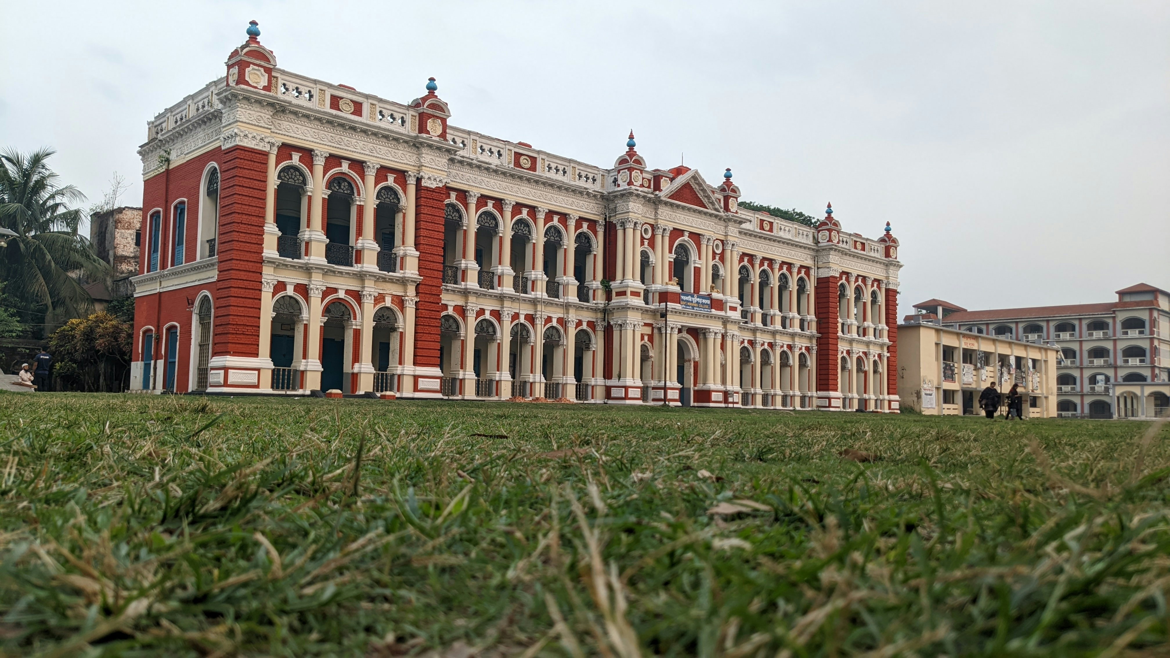 Historic red-brick villa with white arched colonnades sits on a manicured lawn under an overcast sky.