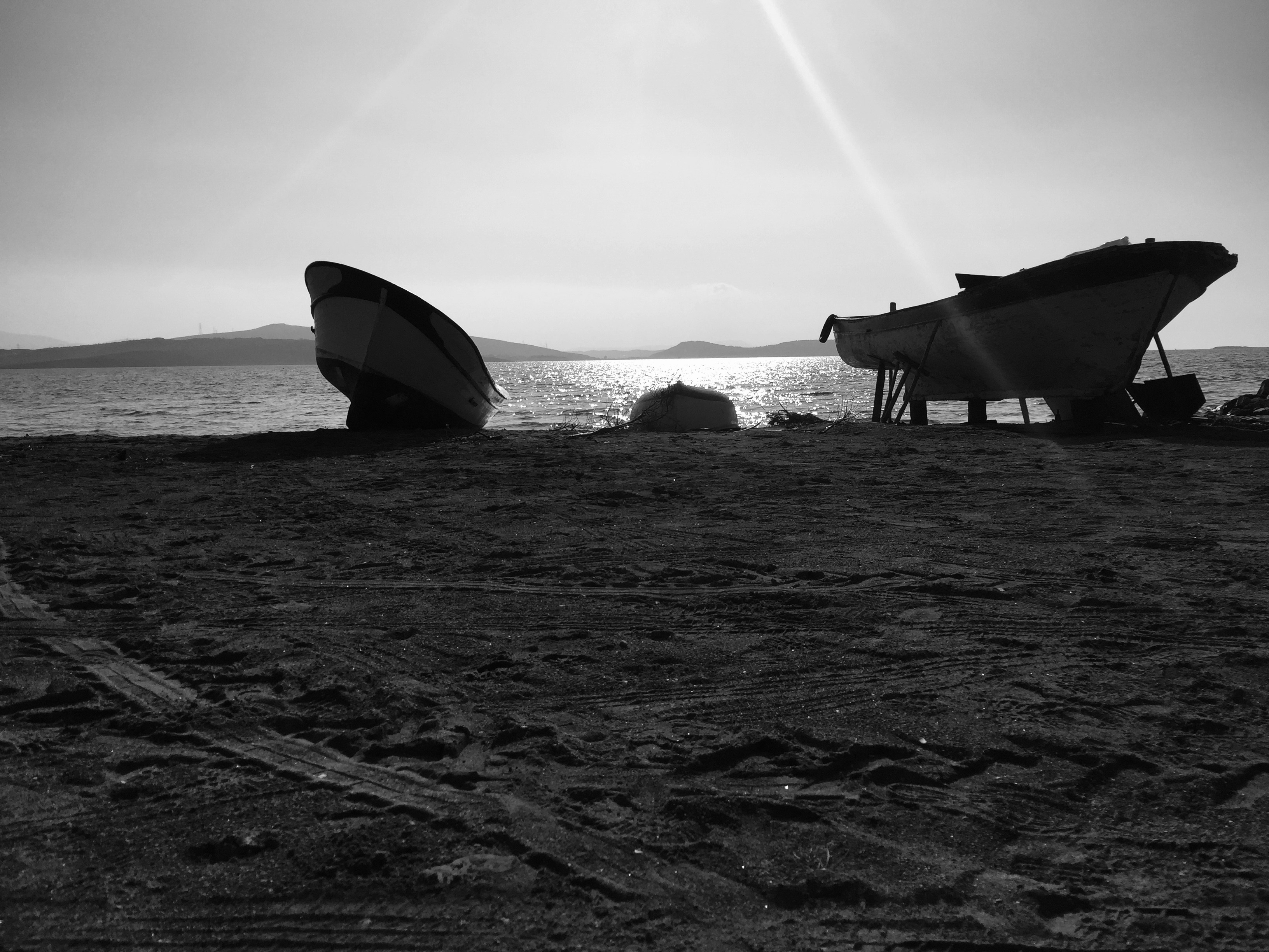Silhouetted boats rest on a sandy shore with a bright sun casting light over calm waters.