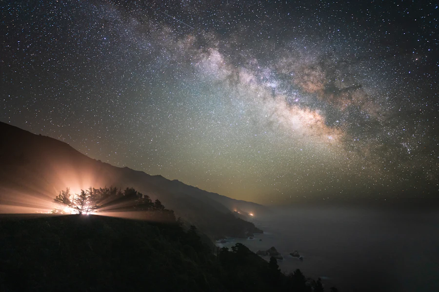 The Milky Way arcs across a star-filled night sky above dark coastal hills and fog. A warm light glows from a small tree on the left, adding contrast to the scene.