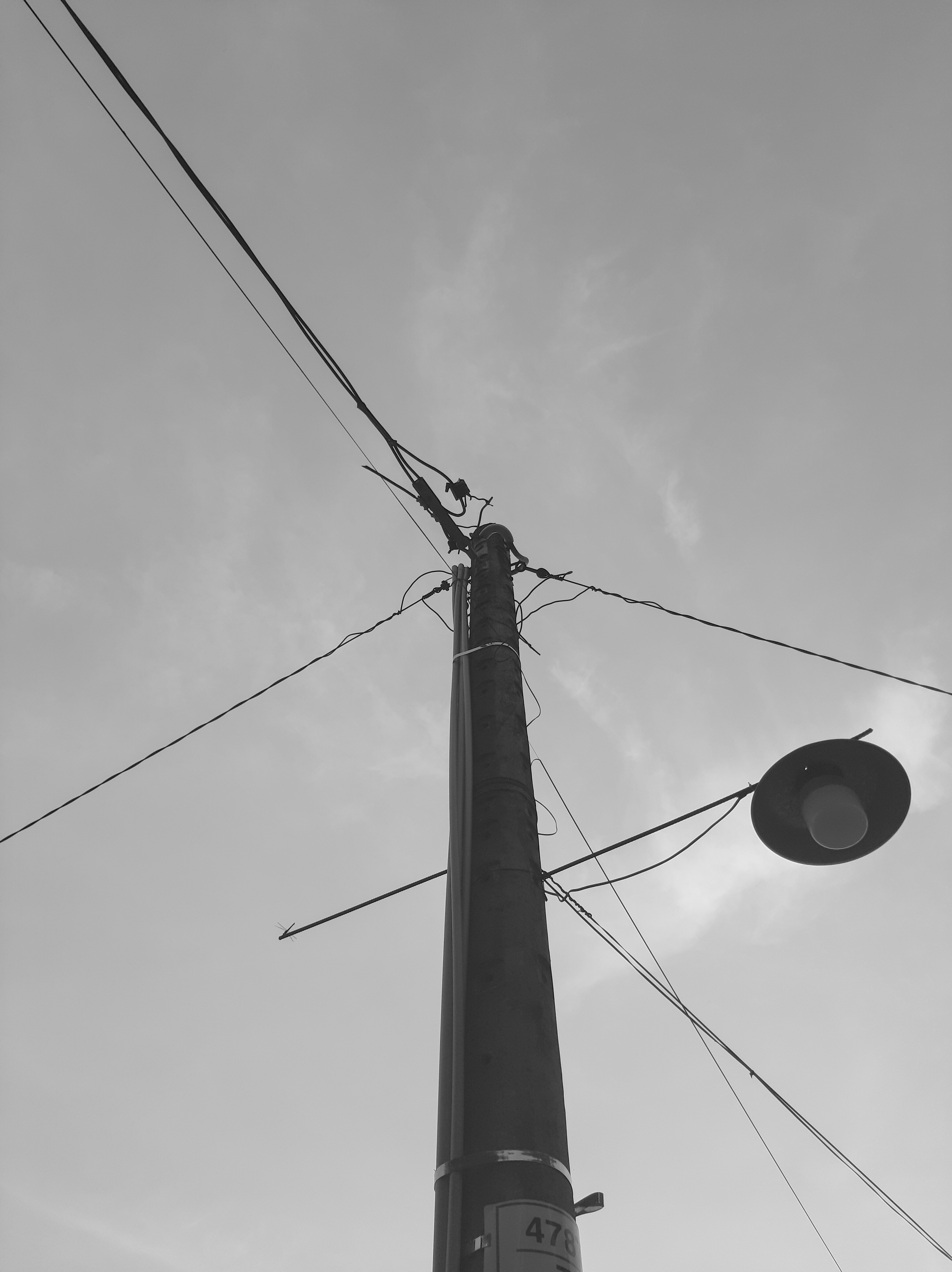 Vertical view of a utility pole with intersecting wires and a streetlight against a cloudy sky.