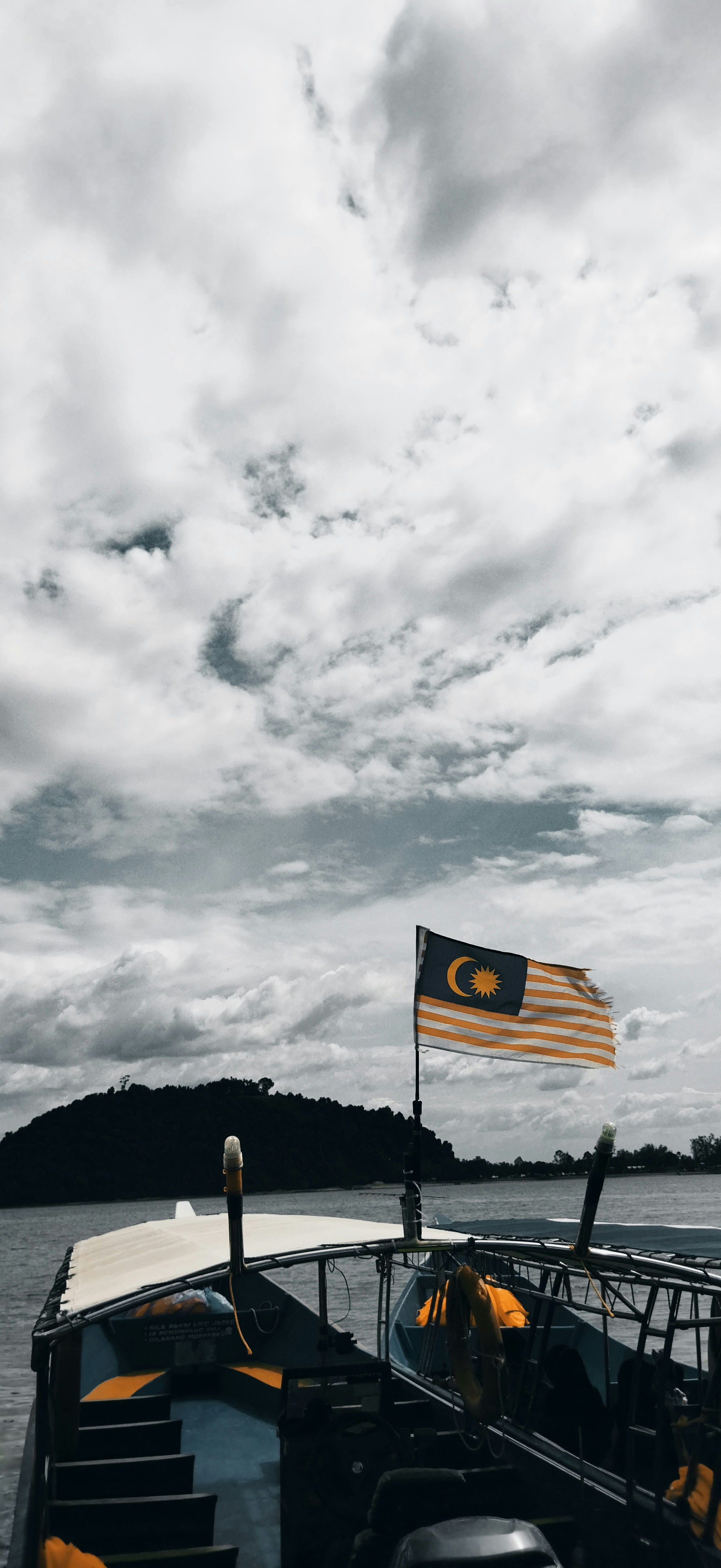 A boat navigating the serene waters of Malaysia, adorned with a flag that symbolizes national pride against a backdrop of dramatic clouds.