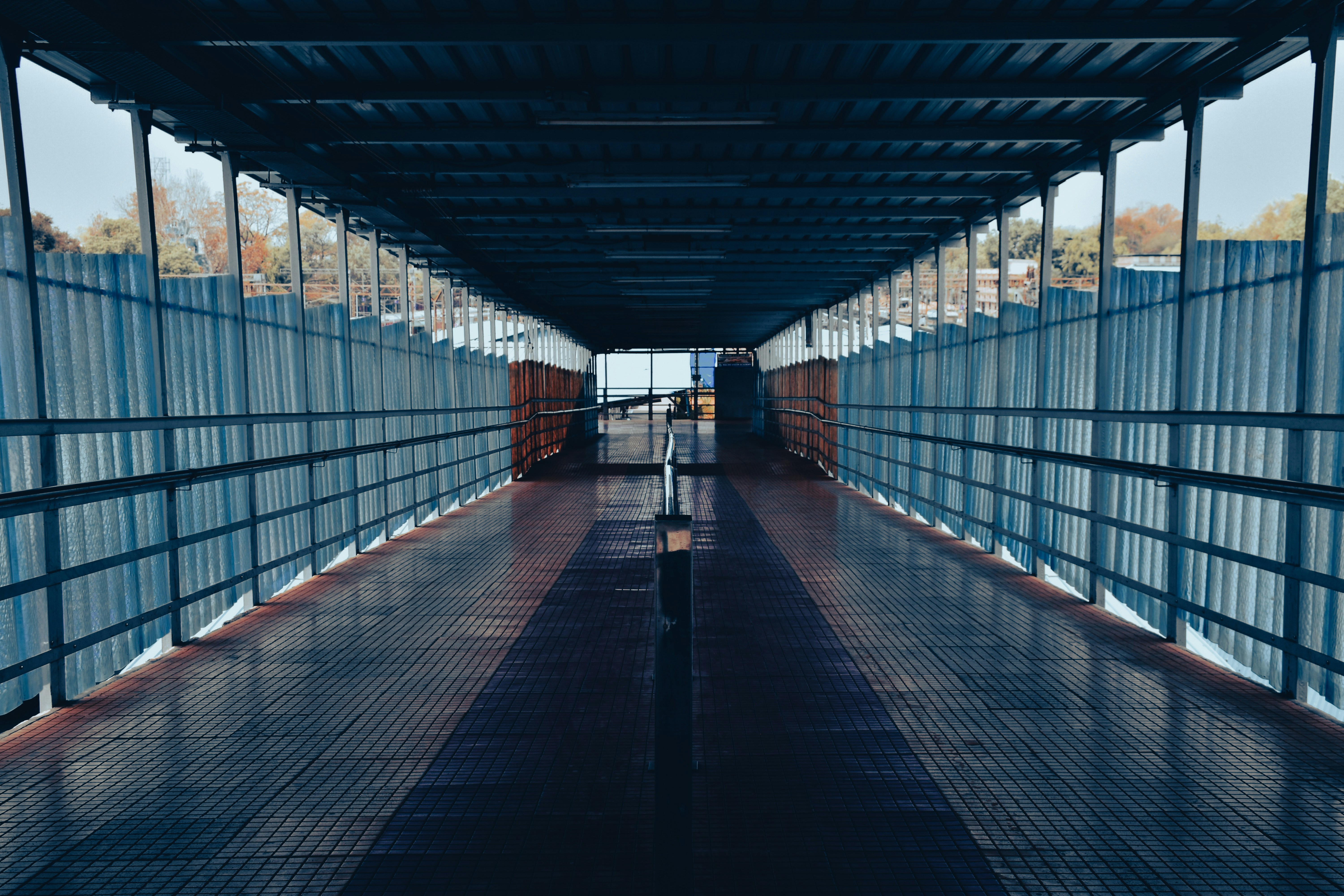 Long, narrow corridor with blue railings and a reflective floor, leading to a bright exit. The structure showcases a blend of light and shadow.