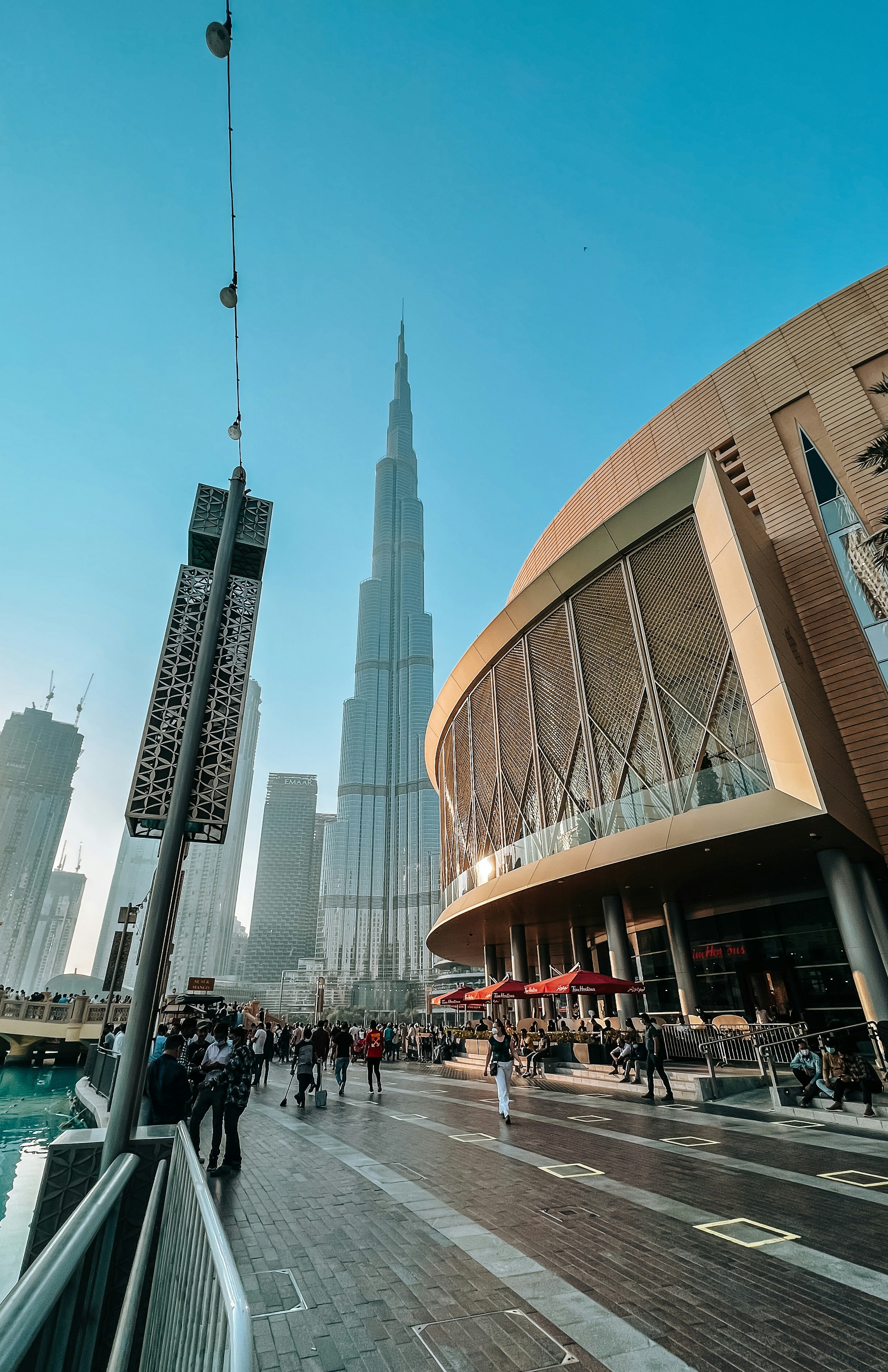 The Burj Khalifa towers majestically over a bustling promenade, with a contemporary structure in the foreground. People stroll leisurely beneath the clear blue sky.