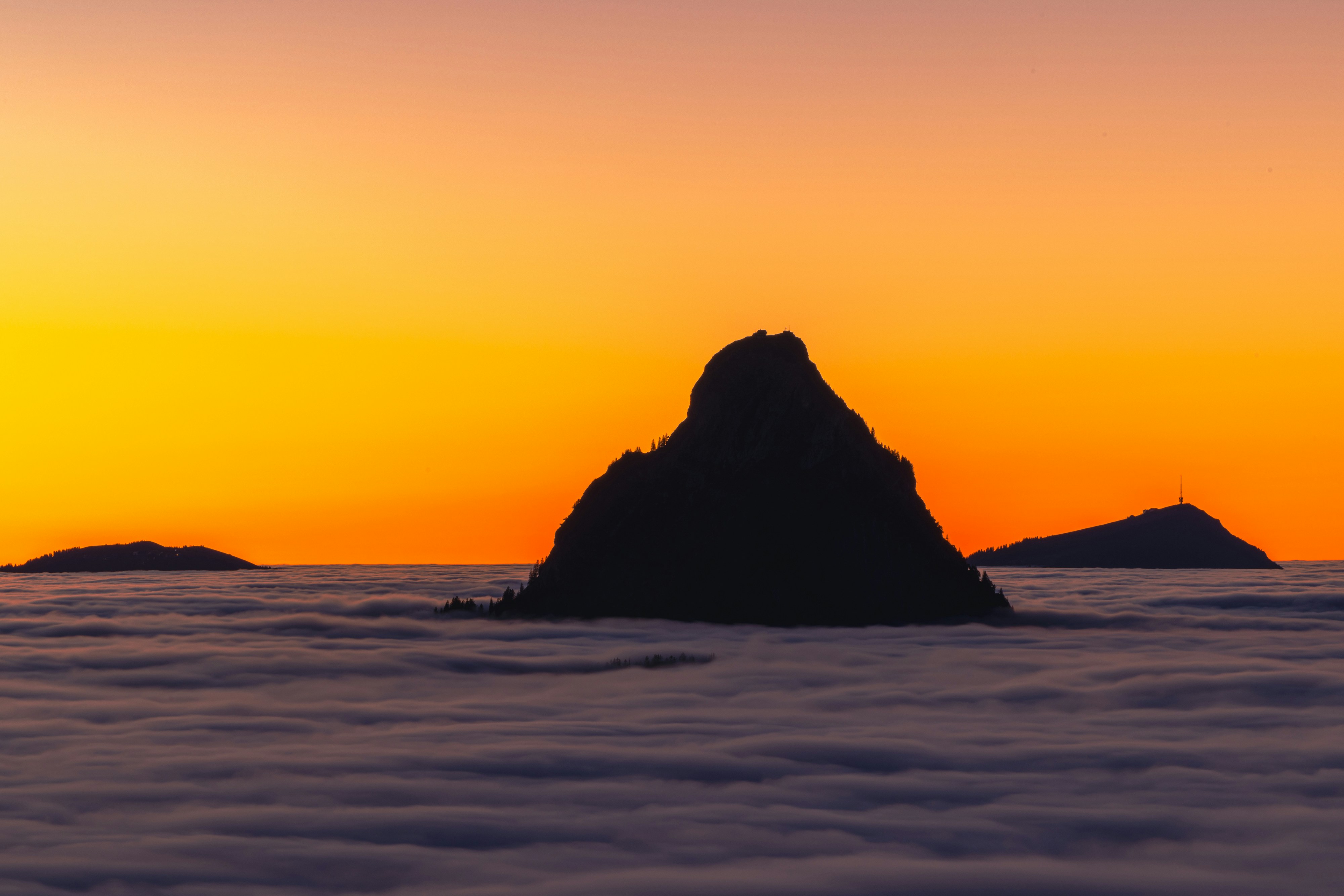 Mountain peaks rise through a dense cloud layer under a vibrant orange sunrise sky.