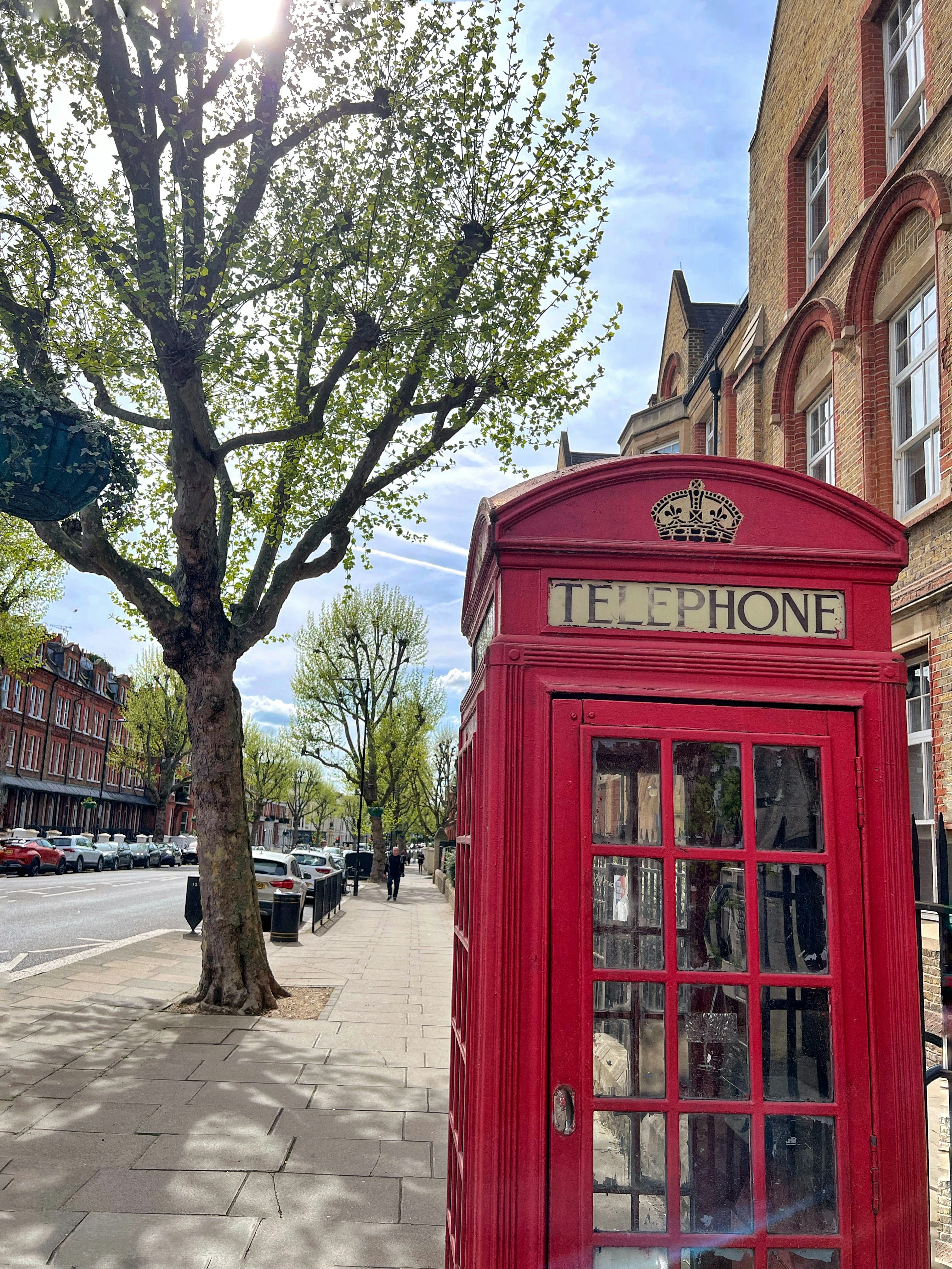 Red Kiosk on a Crooked Street: A London-esque Telegraph Stand Beside Bright Foliage