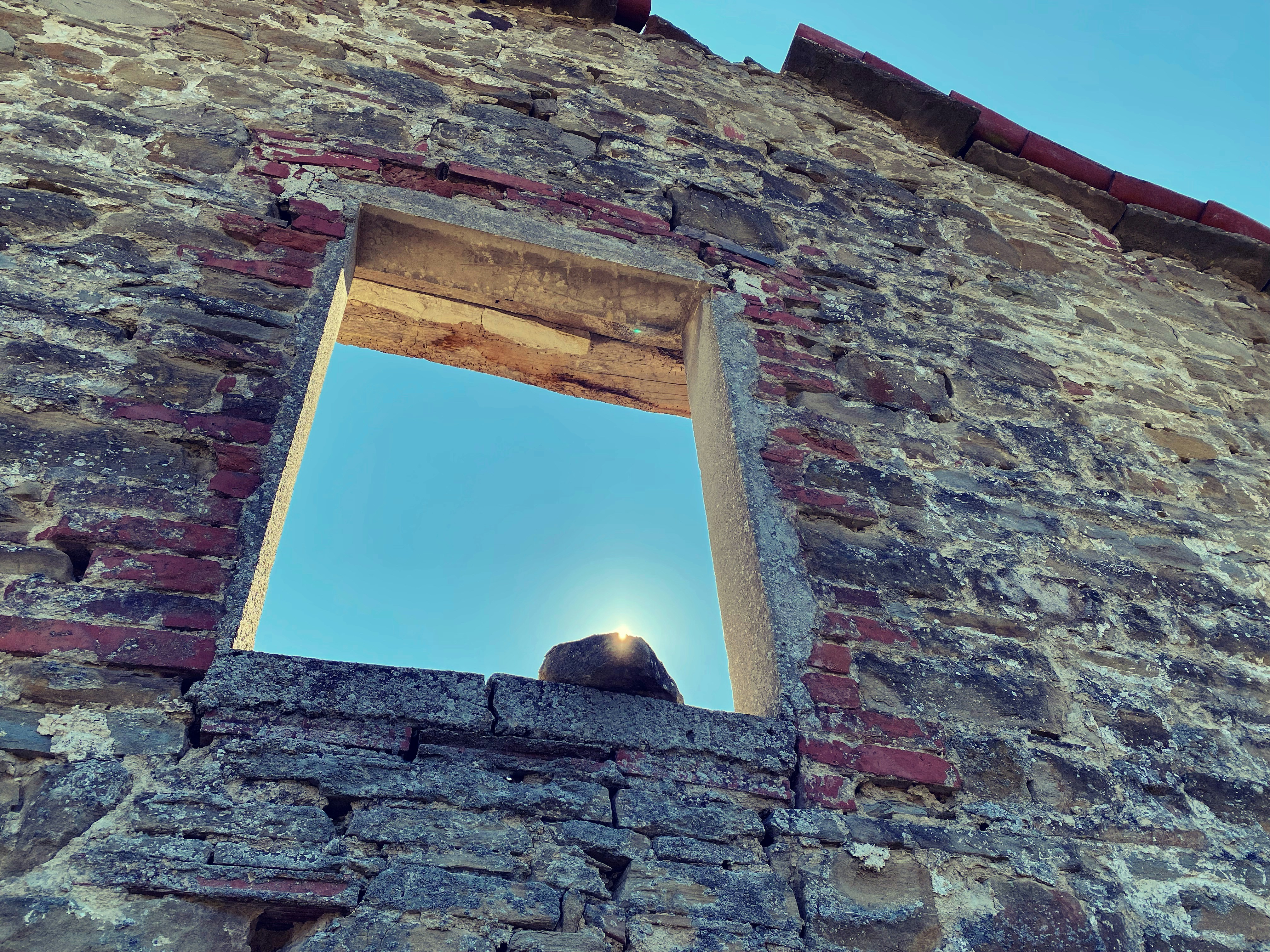 Sunlight streaming through a weathered stone window, illuminating the sky beyond. The rustic texture of the wall contrasts with the bright blue above.