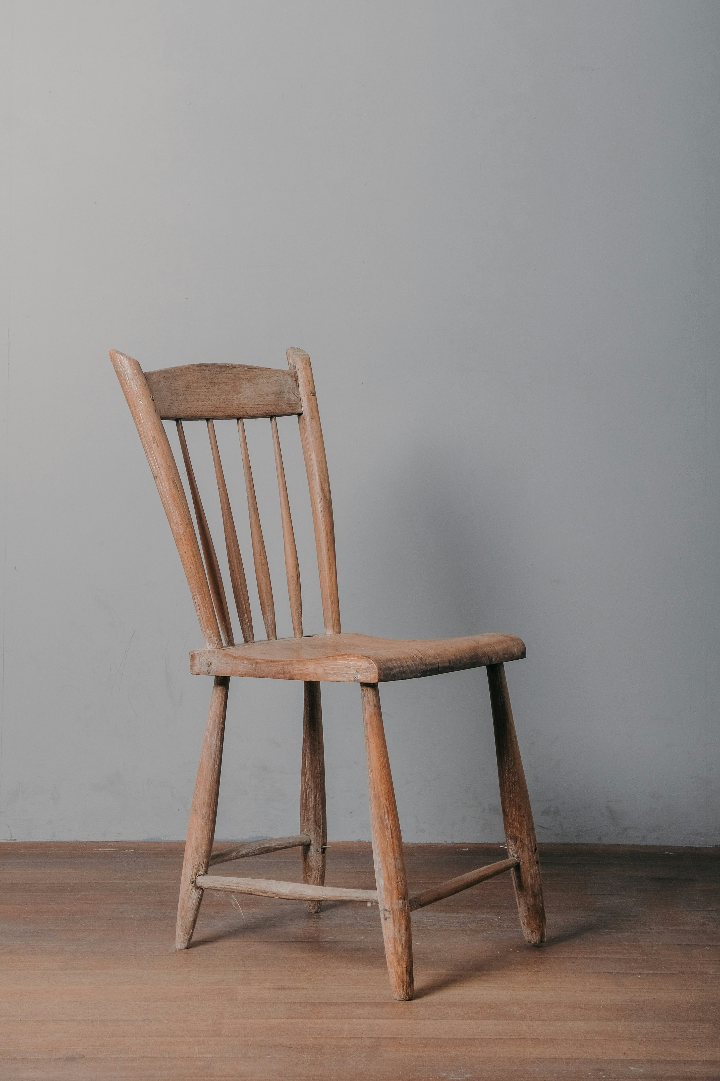 A rustic wooden chair with a minimalist design, set against a muted gray backdrop. The natural textures of the wood stand out prominently.