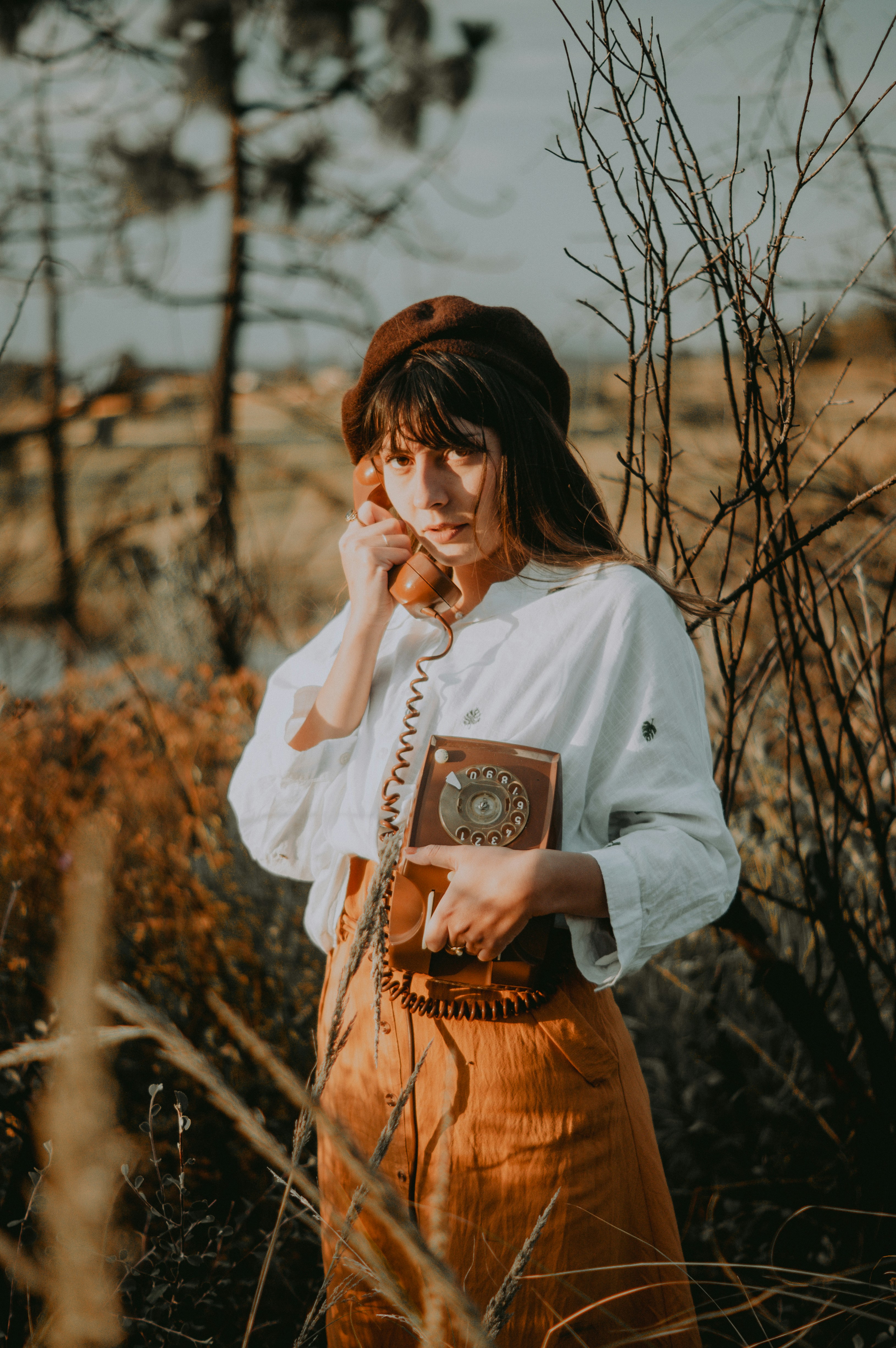 A woman in a white blouse and brown skirt stands in a sunlit field, speaking on a vintage rotary telephone while clutching a retro leather case.