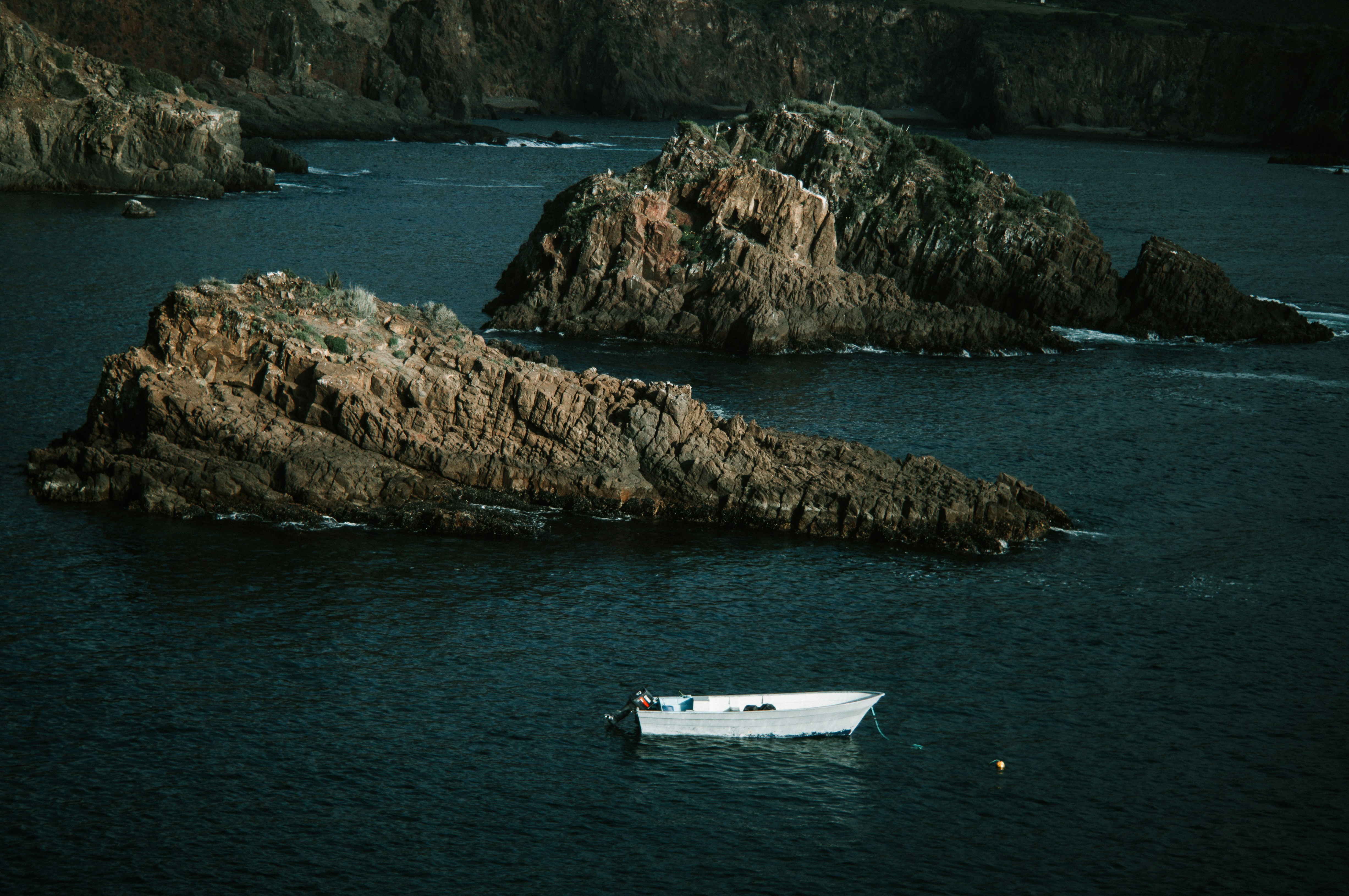 A photograph shows a small white boat resting in calm blue water beside rugged rocky outcrops. The composition emphasizes solitude against the jagged coastline.