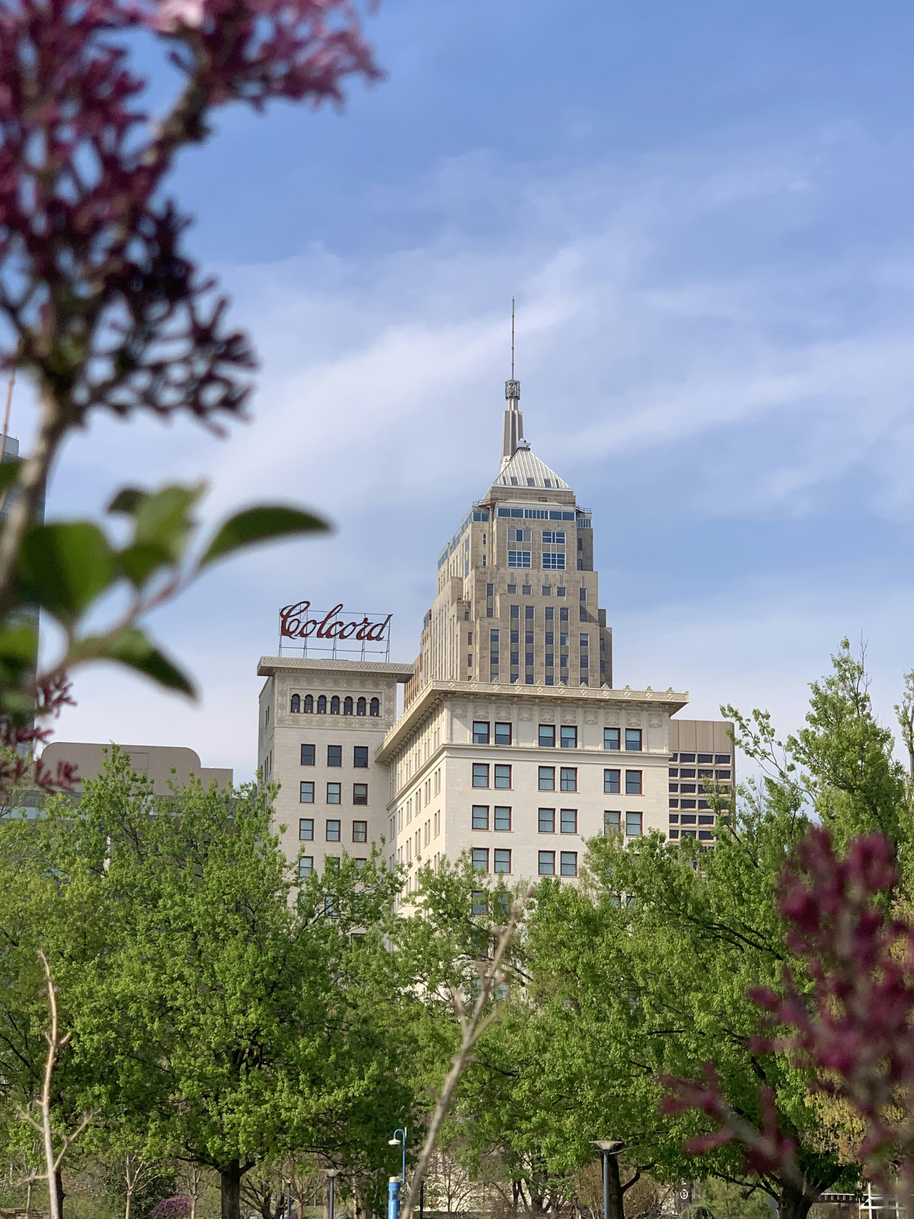 Historic Colcord Tower rises above vibrant greenery and blooming flowers, showcasing its architectural beauty against a clear sky.