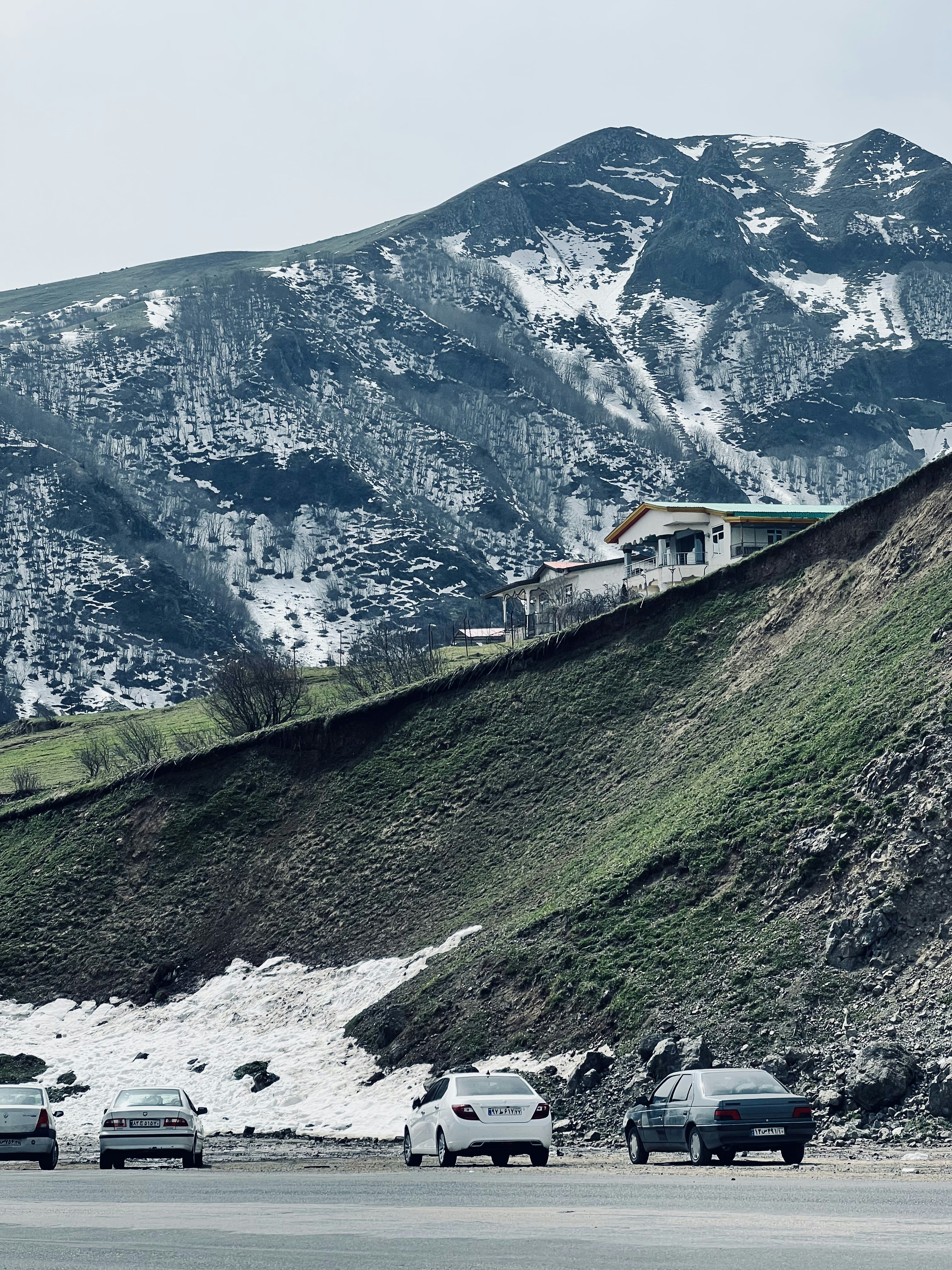 Snow-capped mountains rise behind a green hillside, with parked cars in the foreground. A modest house perches on the slope, showcasing the harmony between human habitation and nature.