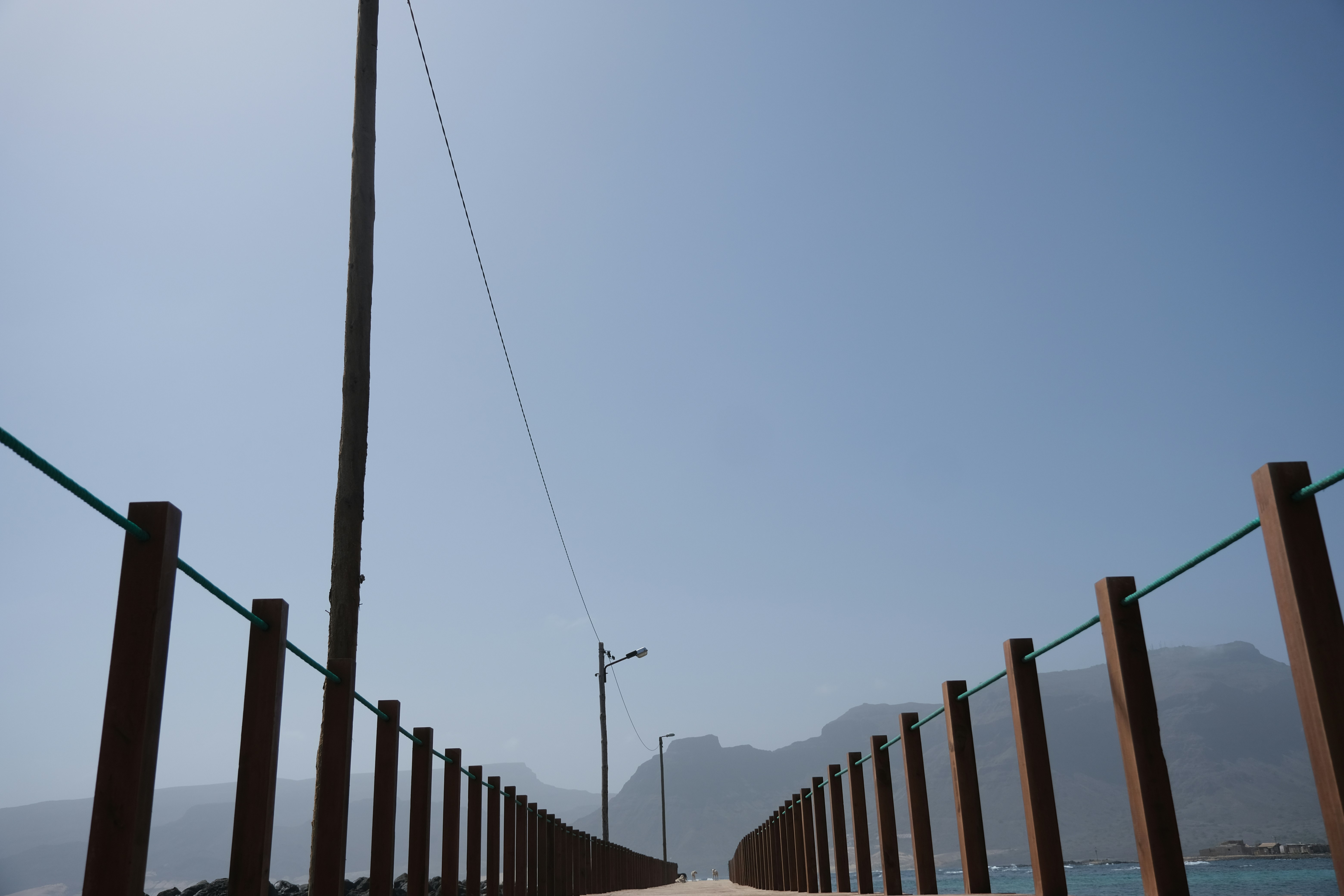 Wooden walkway leading towards distant mountains under a clear blue sky, with a lone street lamp adding a touch of solitude.