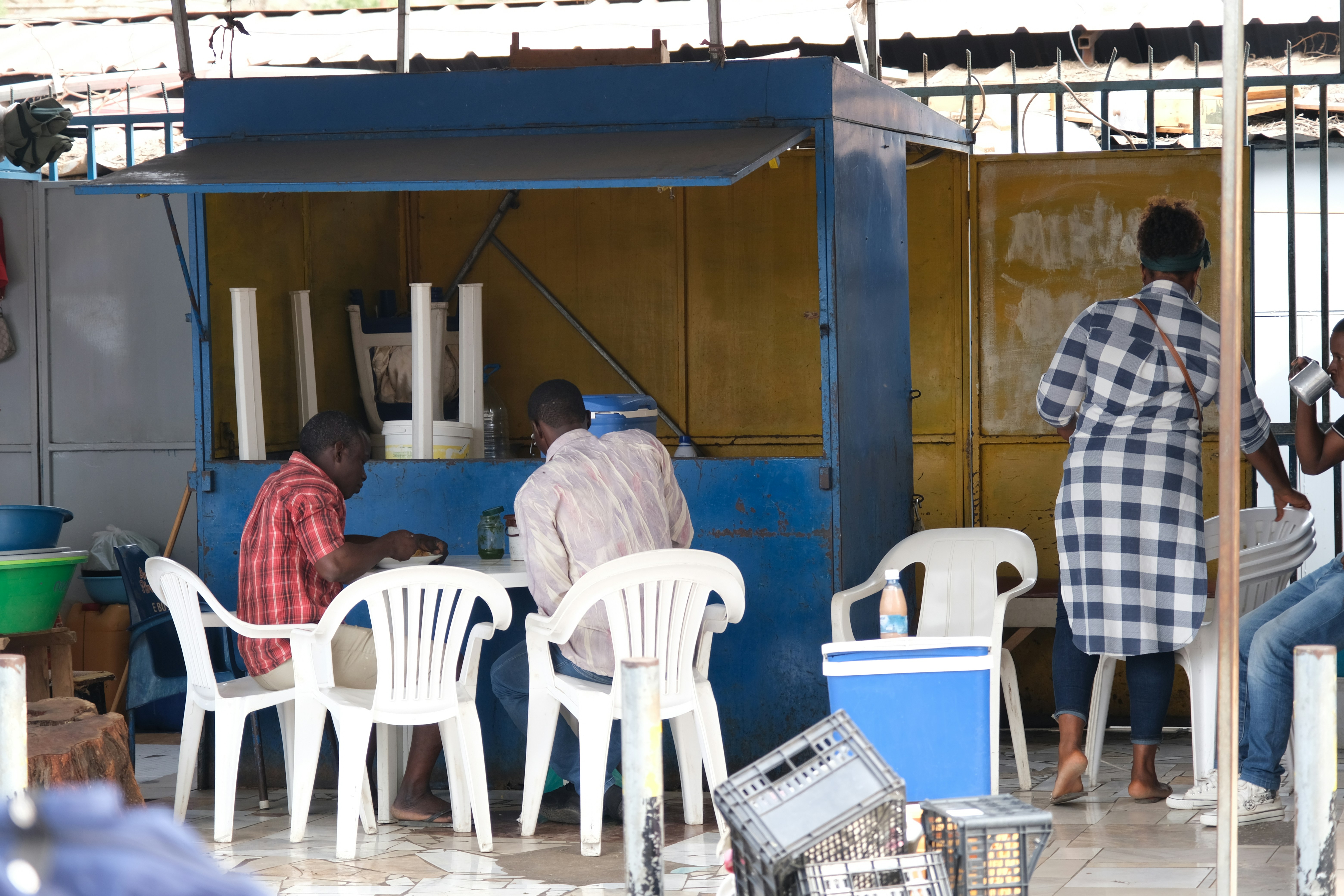 Two men engaged in conversation at a market stall, surrounded by colorful containers and a busy atmosphere. A woman in a checkered outfit walks by, adding to the lively scene.