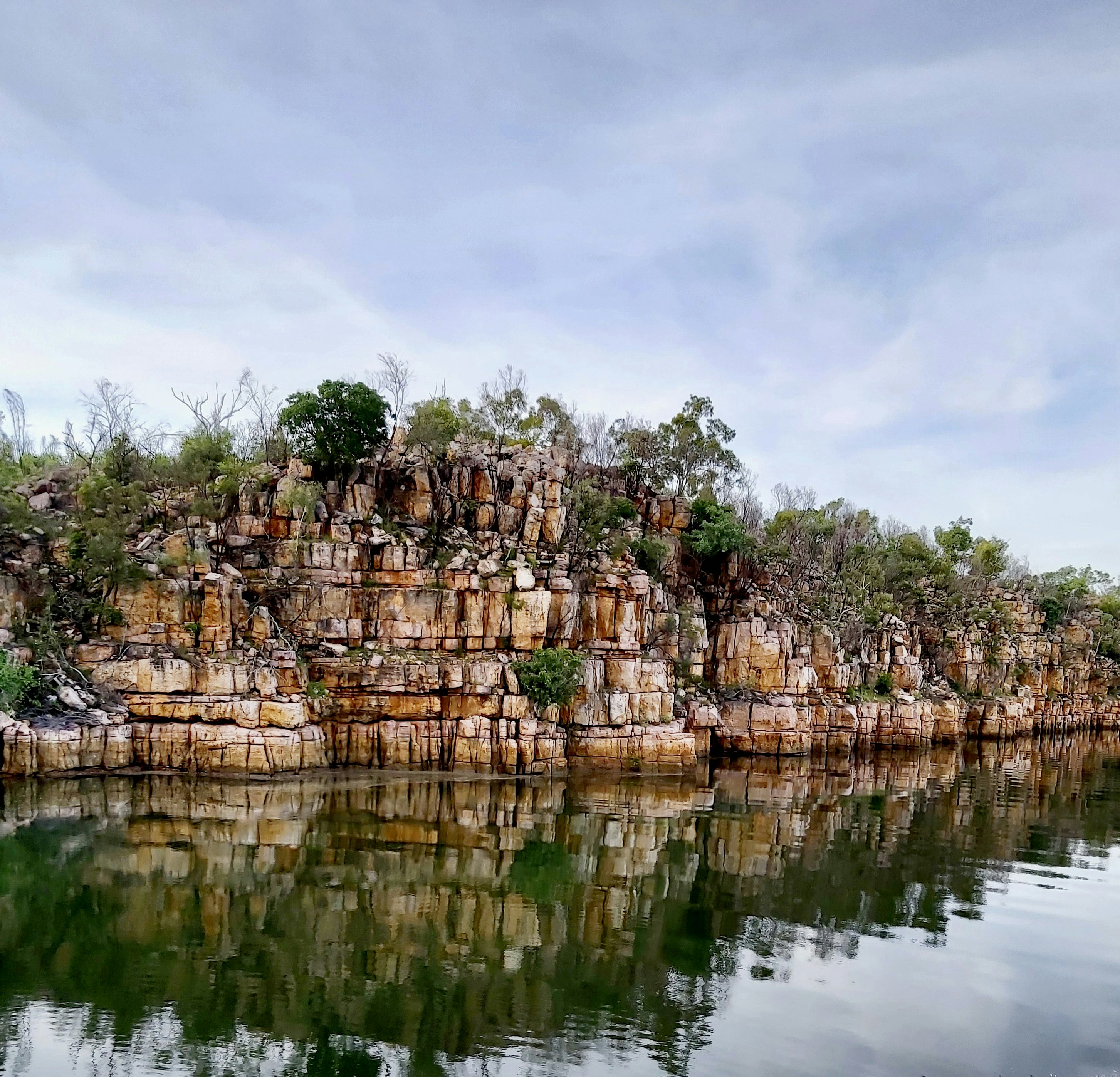 Lush green vegetation crowns rugged cliffs reflecting in still waters, capturing the serene beauty of nature's artistry.