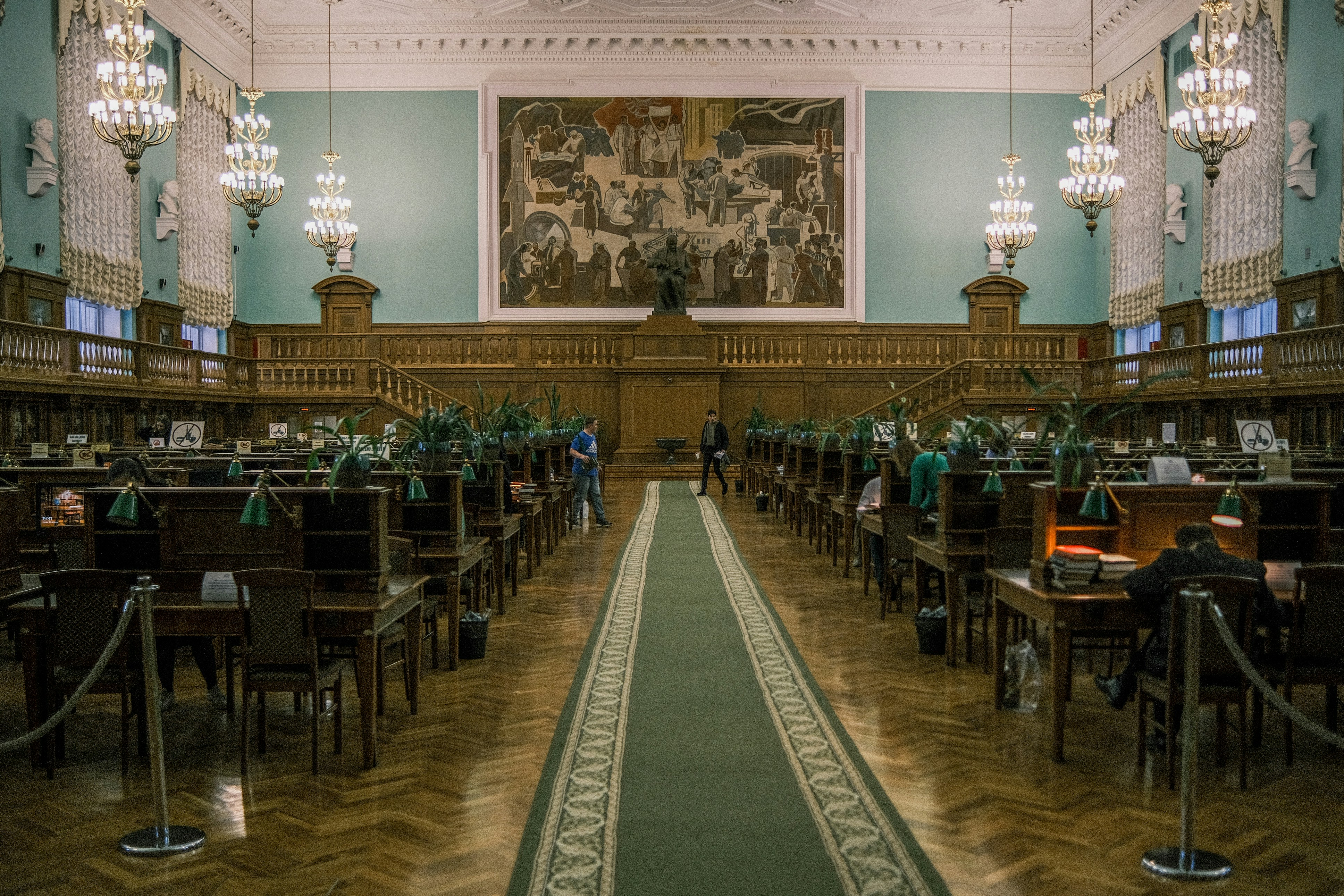 Ornate library hall with rows of wooden desks, green lamps, and a large mural on the back wall.