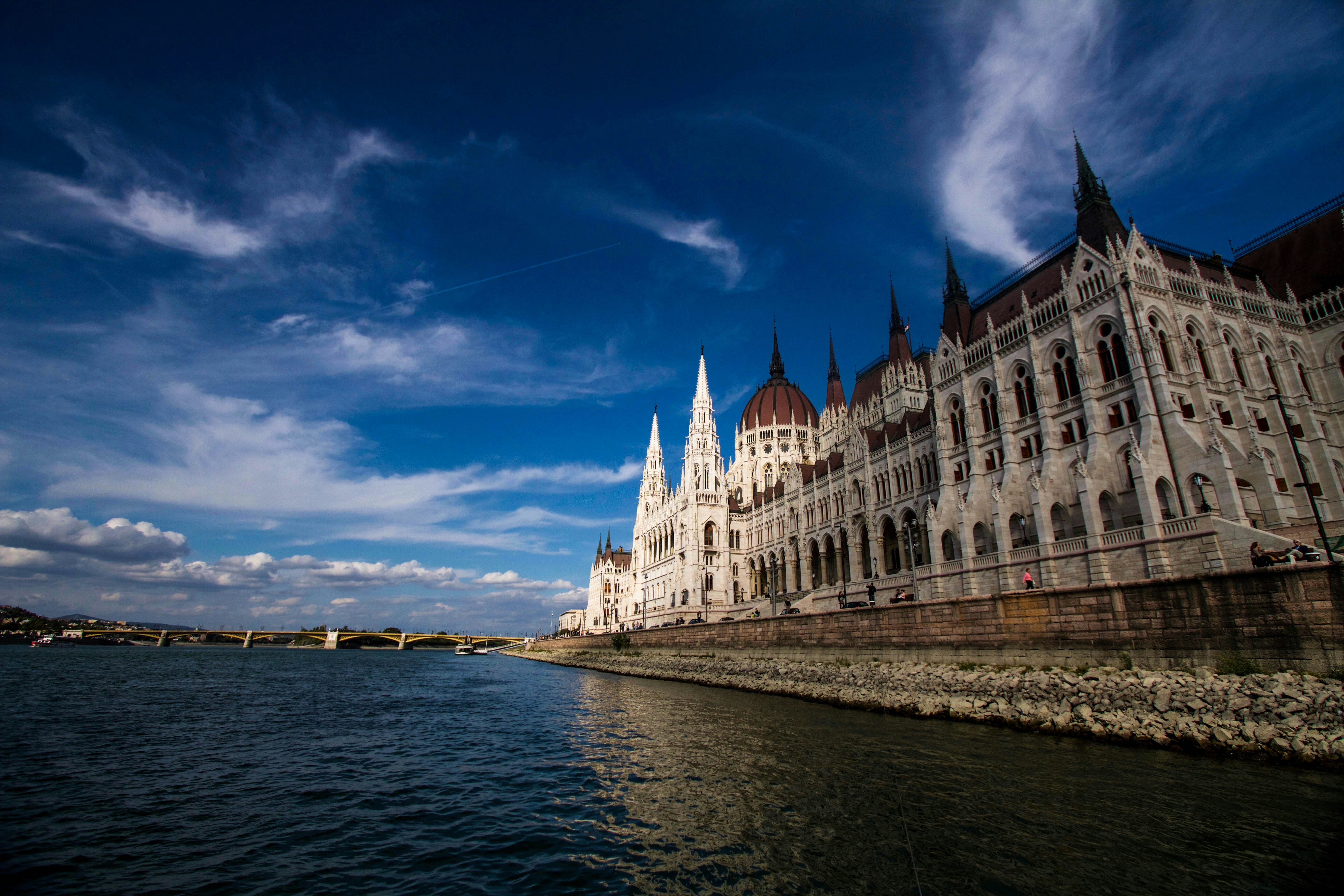 The ornate architecture of the Parliament building rises majestically along the riverbank, framed by a vibrant sky. A serene water reflection enhances the scene.