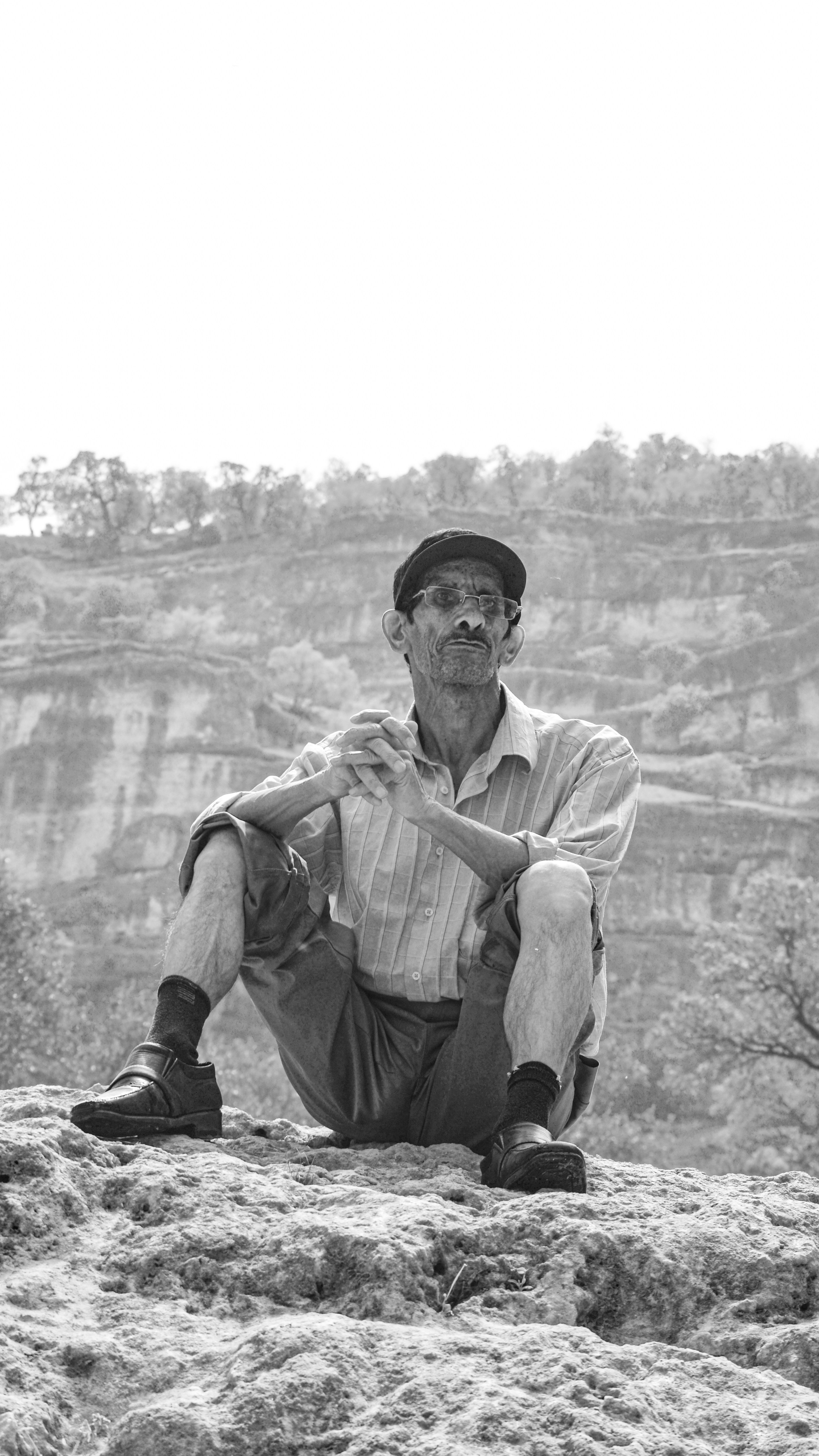 Elderly man seated on a rocky outcrop, gazing thoughtfully into the distance, surrounded by a rugged landscape.