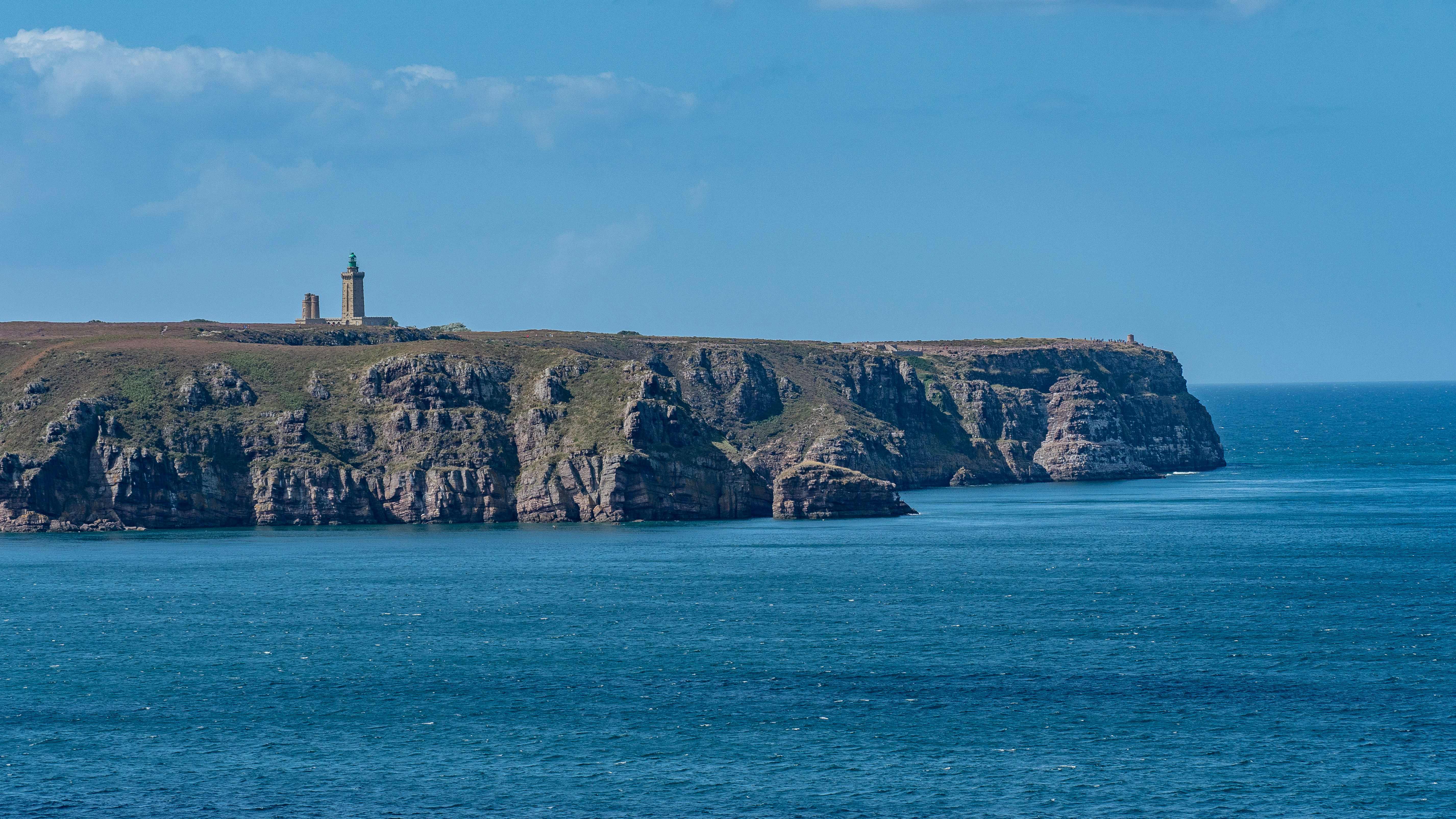 Lighthouse perched on rugged cliffs overlooking a serene sea under a clear blue sky.