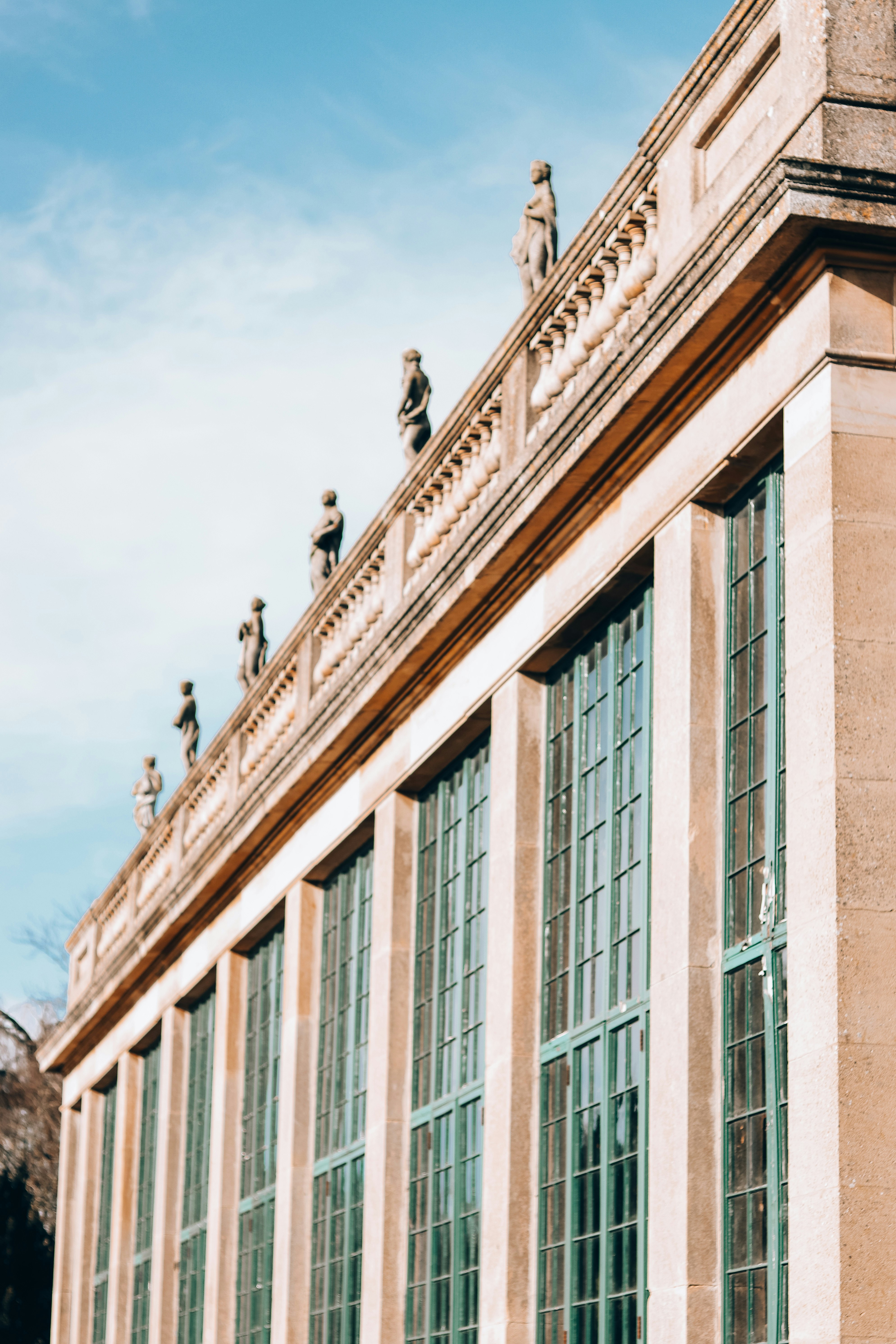 Architectural detail of a historic building featuring statues along the rooftop, framed by large glass windows. The sunlight enhances the textures of the stone and glass.