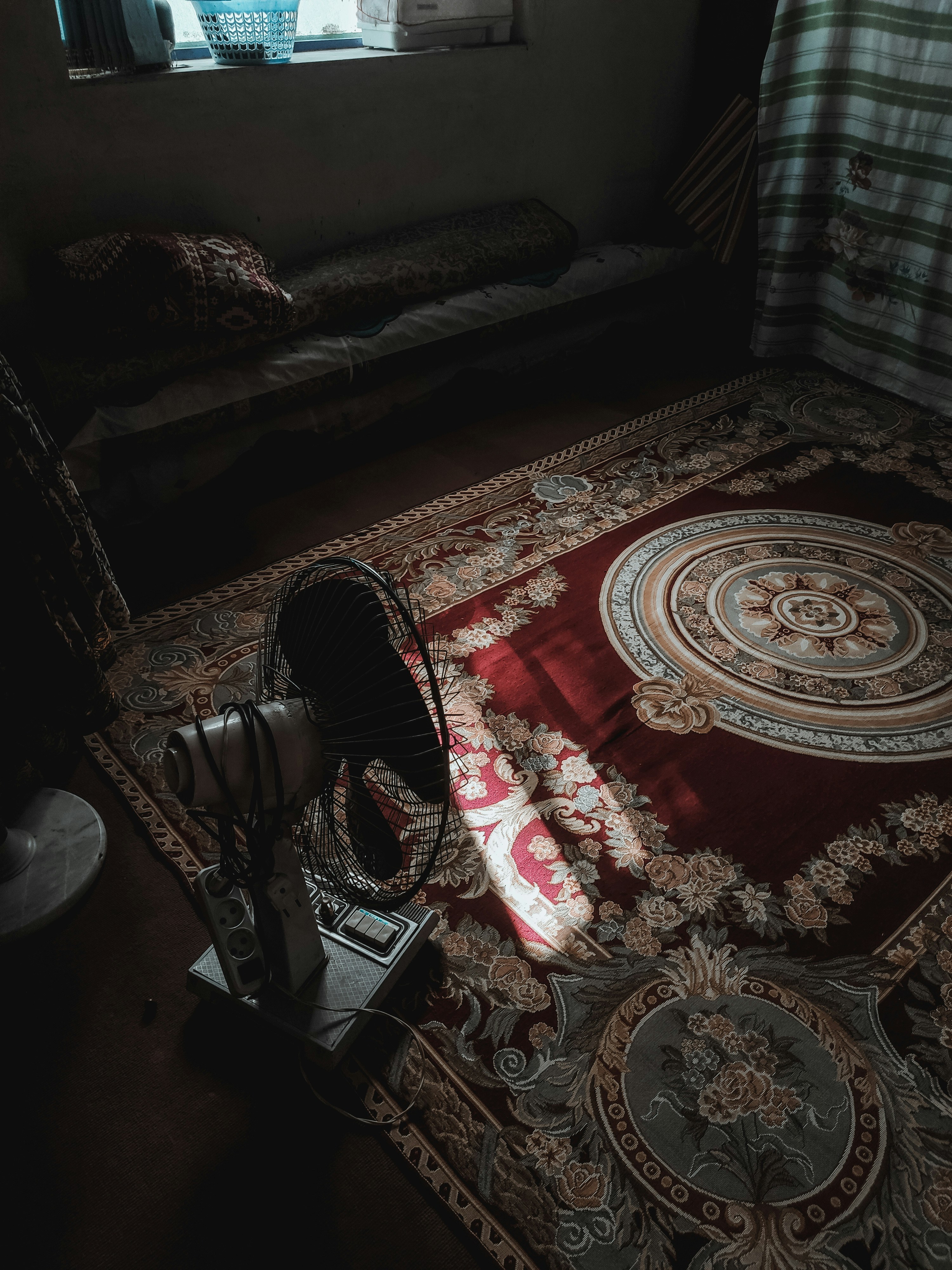 A vintage fan stands prominently on an ornate carpet in a dimly lit room, casting shadows and highlighting the intricate patterns. Sunlight filters through a window, adding warmth to the scene.