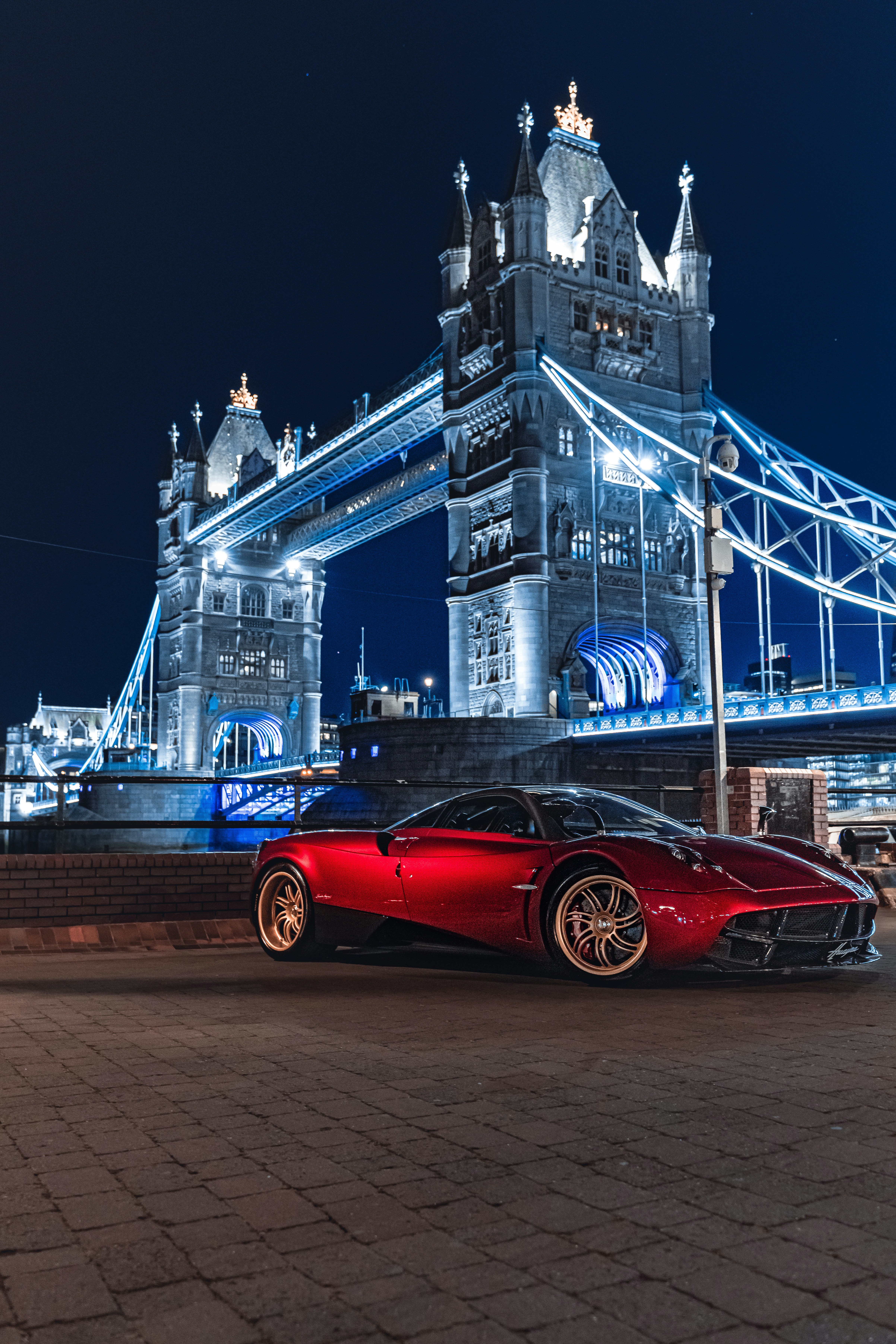 Red sports car sits on cobblestones beneath Tower Bridge lit in blue. The night scene emphasizes contrast between the vehicle and illuminated architectural backdrop.