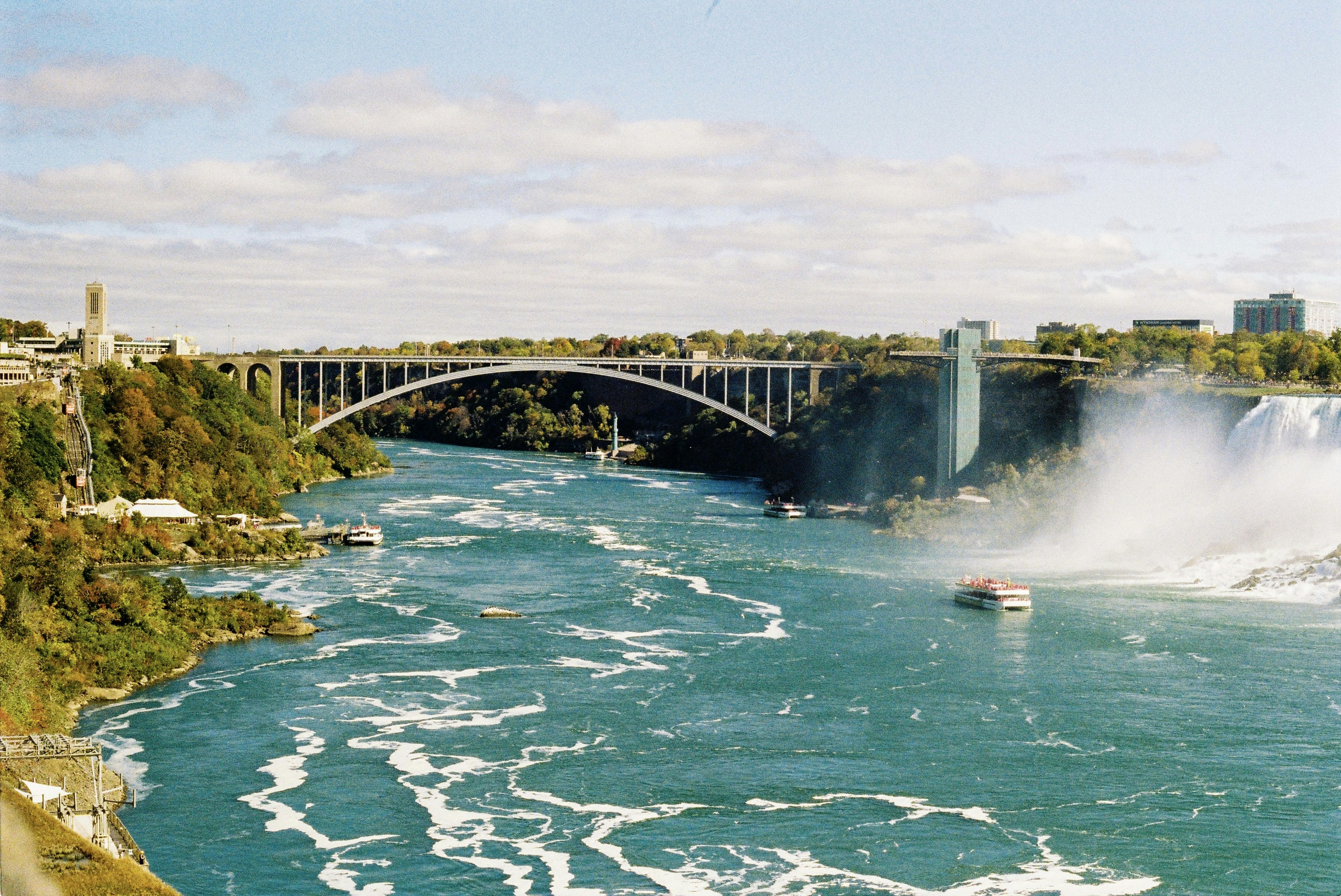 Niagara River flowing beneath a grand arch bridge, with mist rising from the nearby waterfall. A boat navigates the turbulent waters.