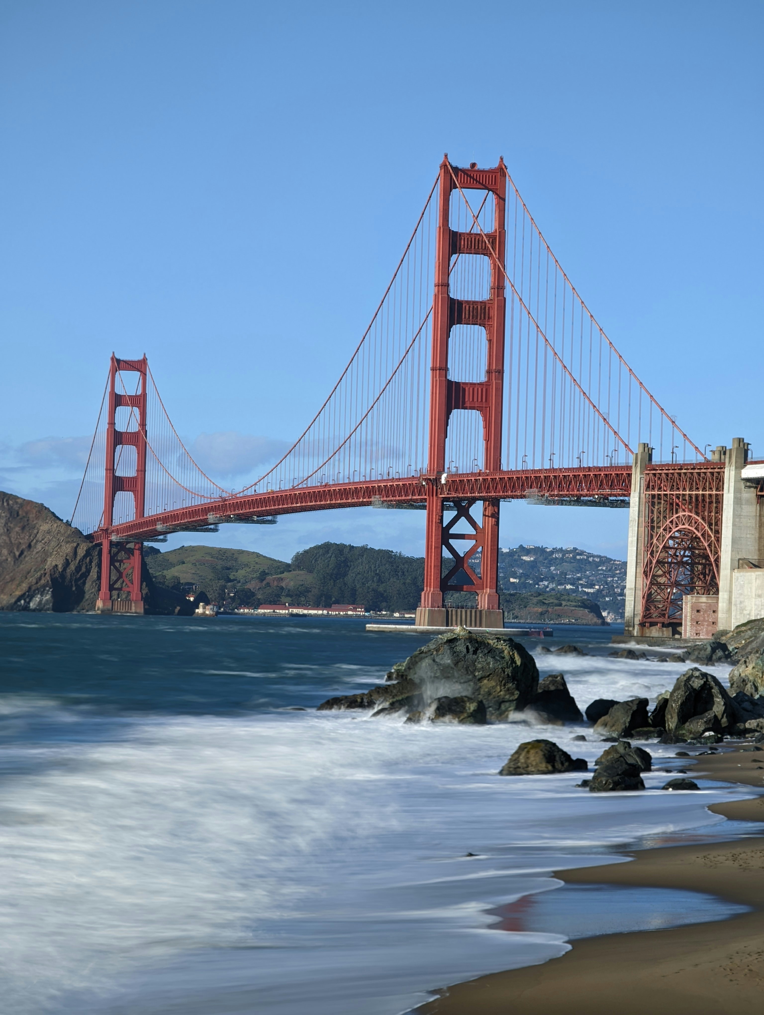 Golden Gate Bridge towering over the coastline, with waves lapping at the rocky shore beneath a clear blue sky.