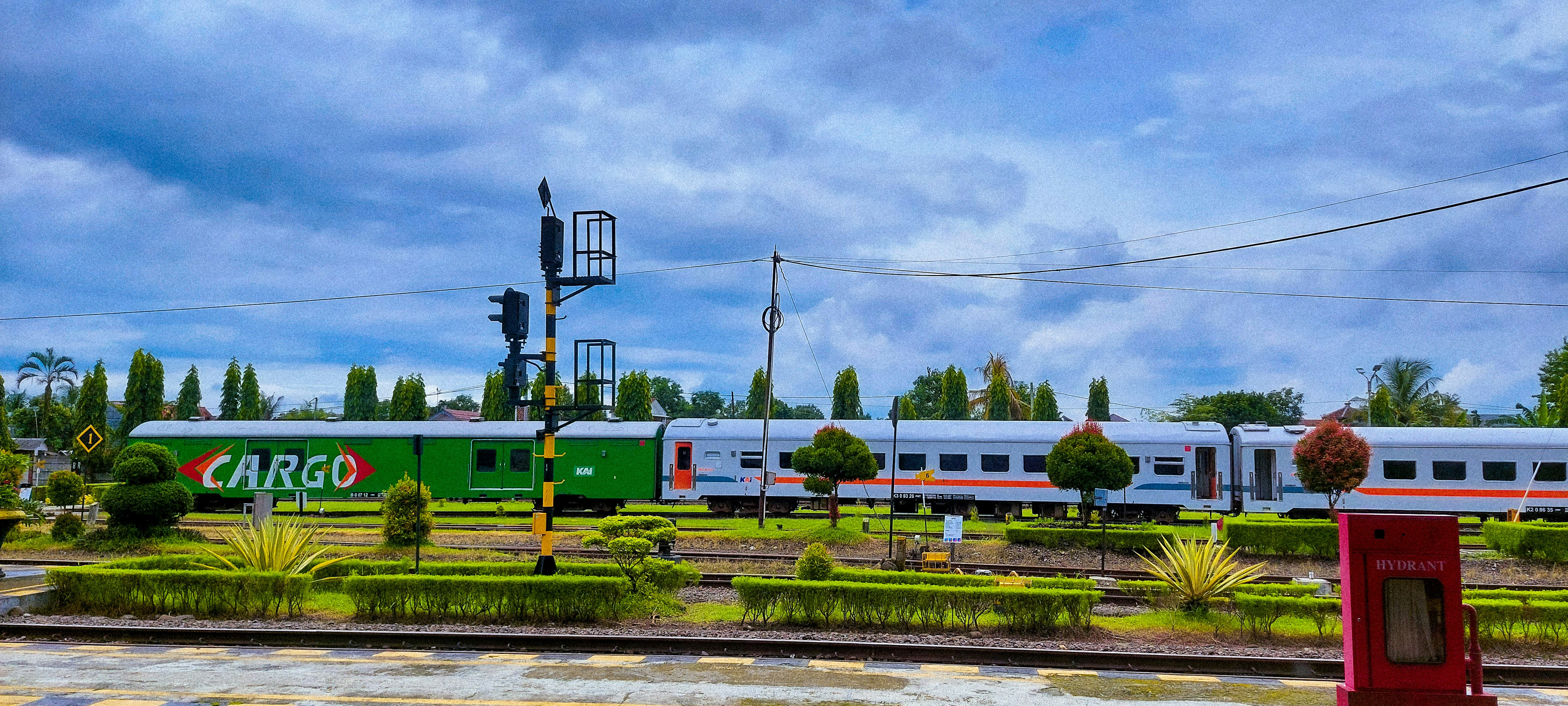 Cargo train passing through a lush, landscaped station under a cloudy sky. The scene captures the essence of rail transport amidst nature.