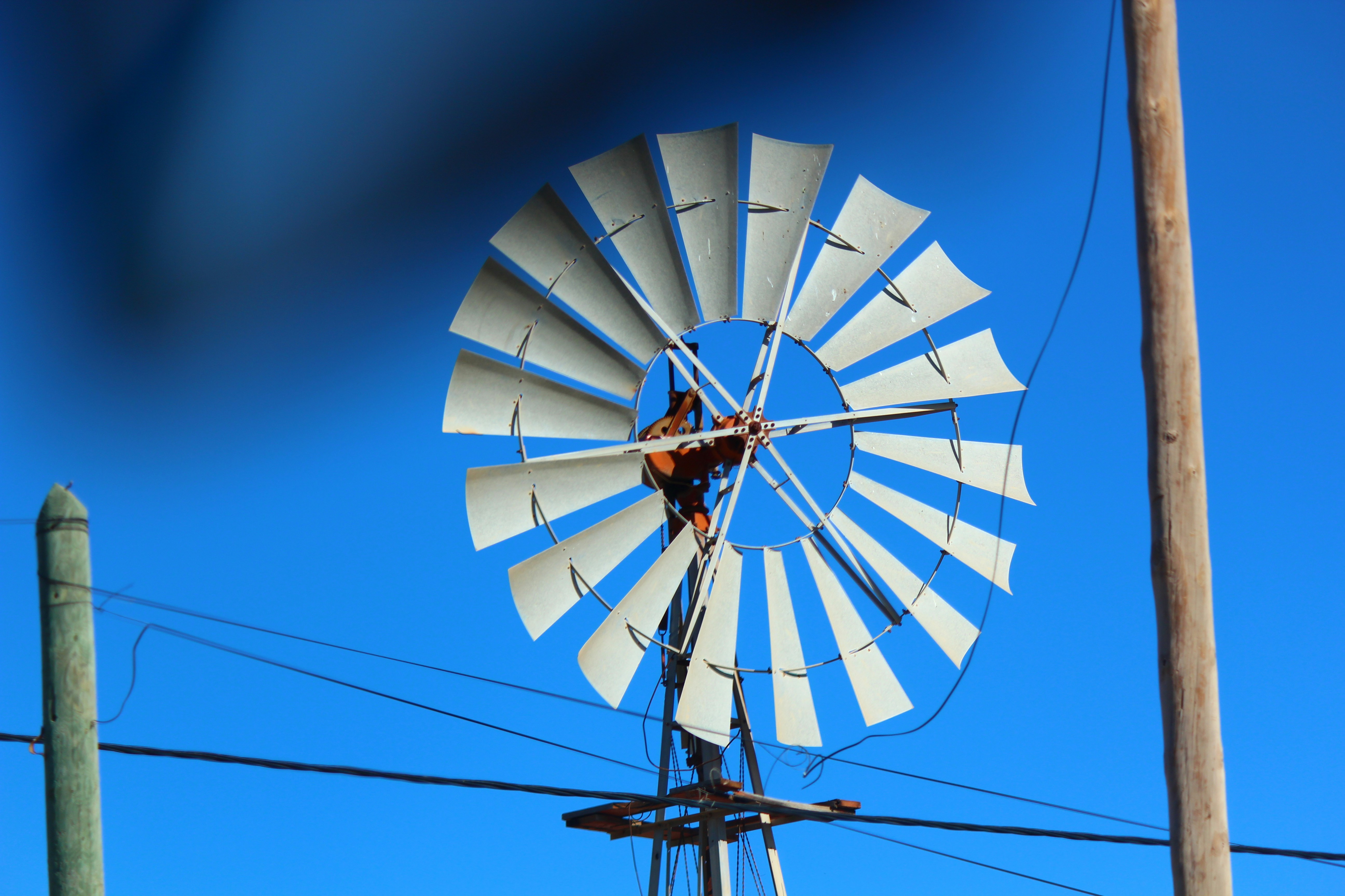A vintage windmill stands tall against a clear blue sky, its blades gracefully spinning in the breeze, surrounded by utility poles and wires.