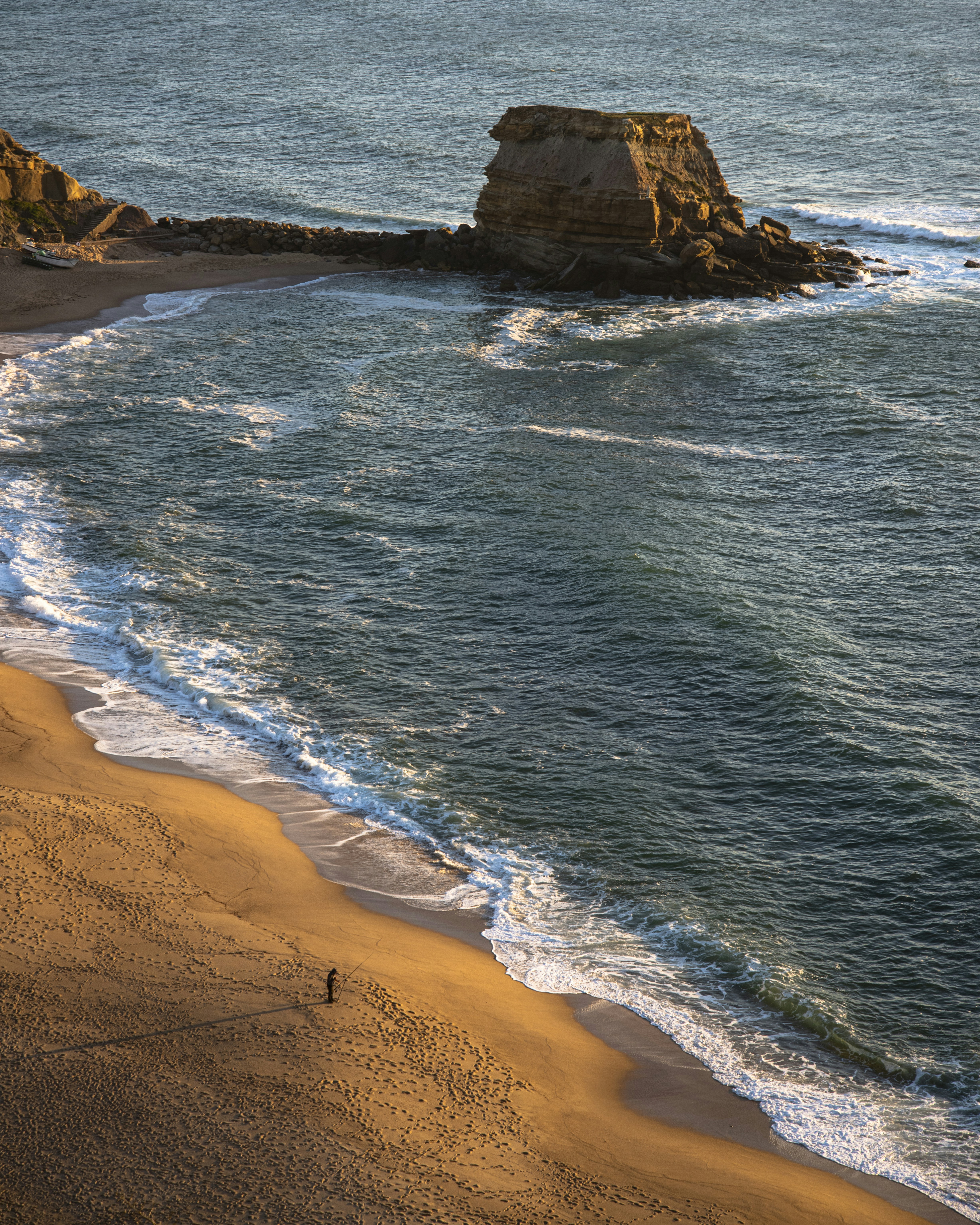 A lone figure stands on a sandy beach, gazing at the gentle waves lapping against a rocky outcrop. The scene captures the tranquility of nature at dusk.