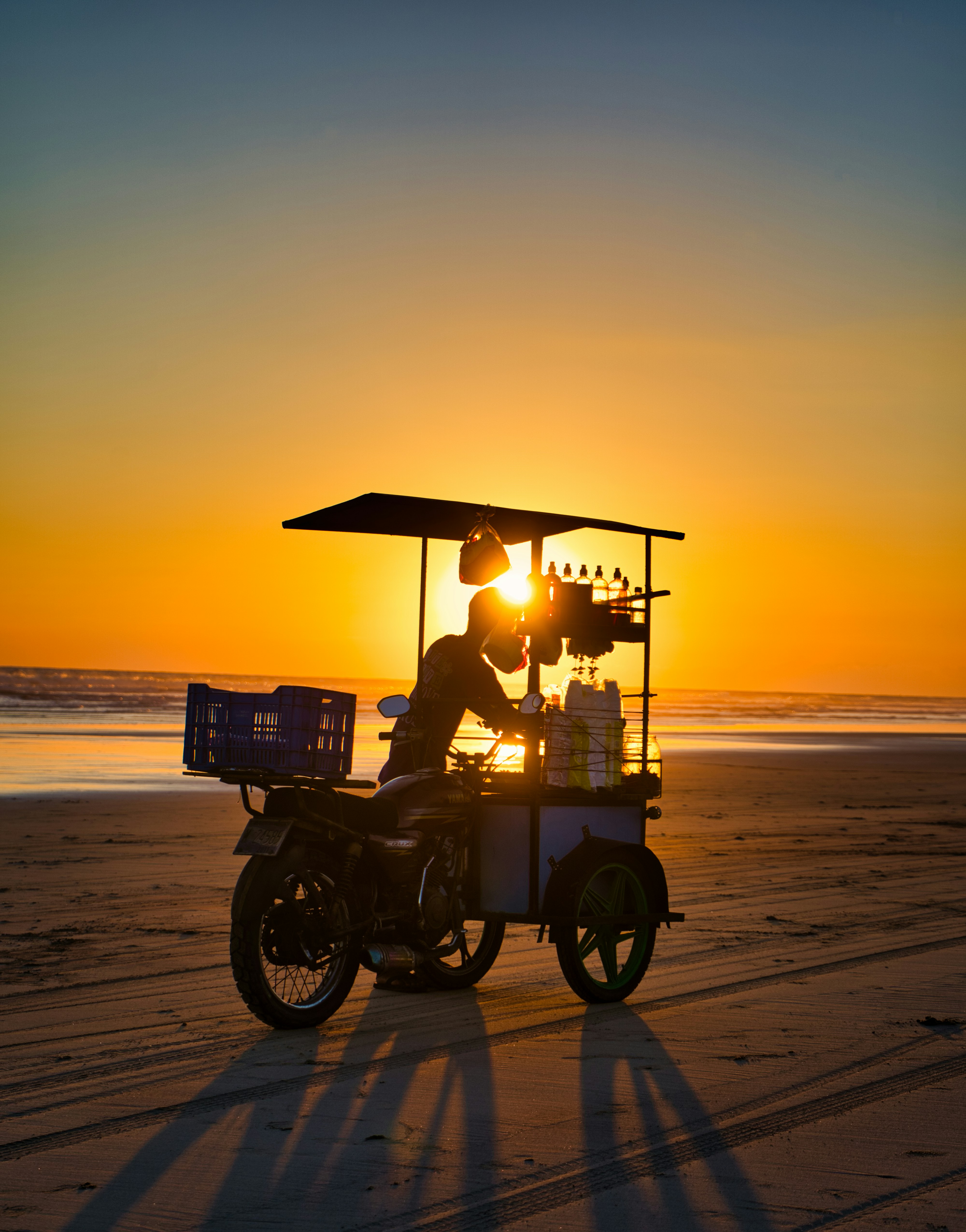 Vendor preparing drinks at a beachside cart as the sun sets on the horizon, casting long shadows on the sand.