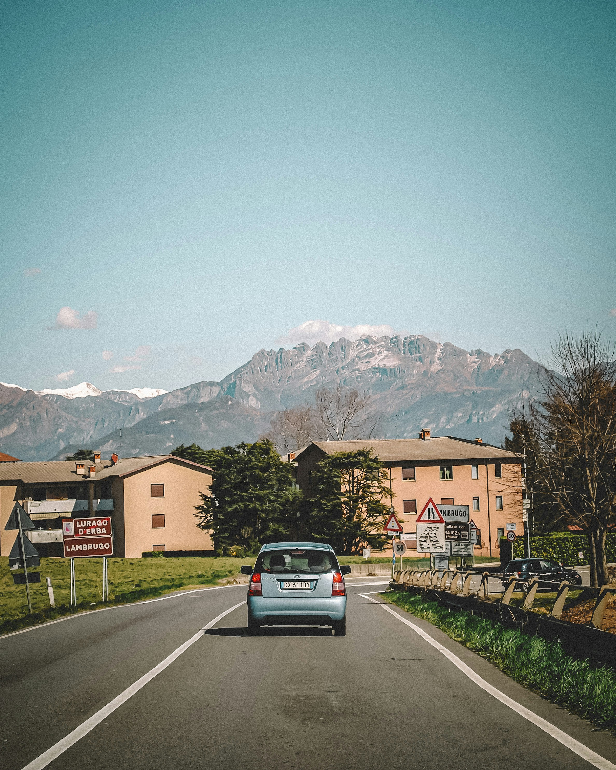 Car driving on a road flanked by buildings with snow-capped mountains in the distance under a clear blue sky.