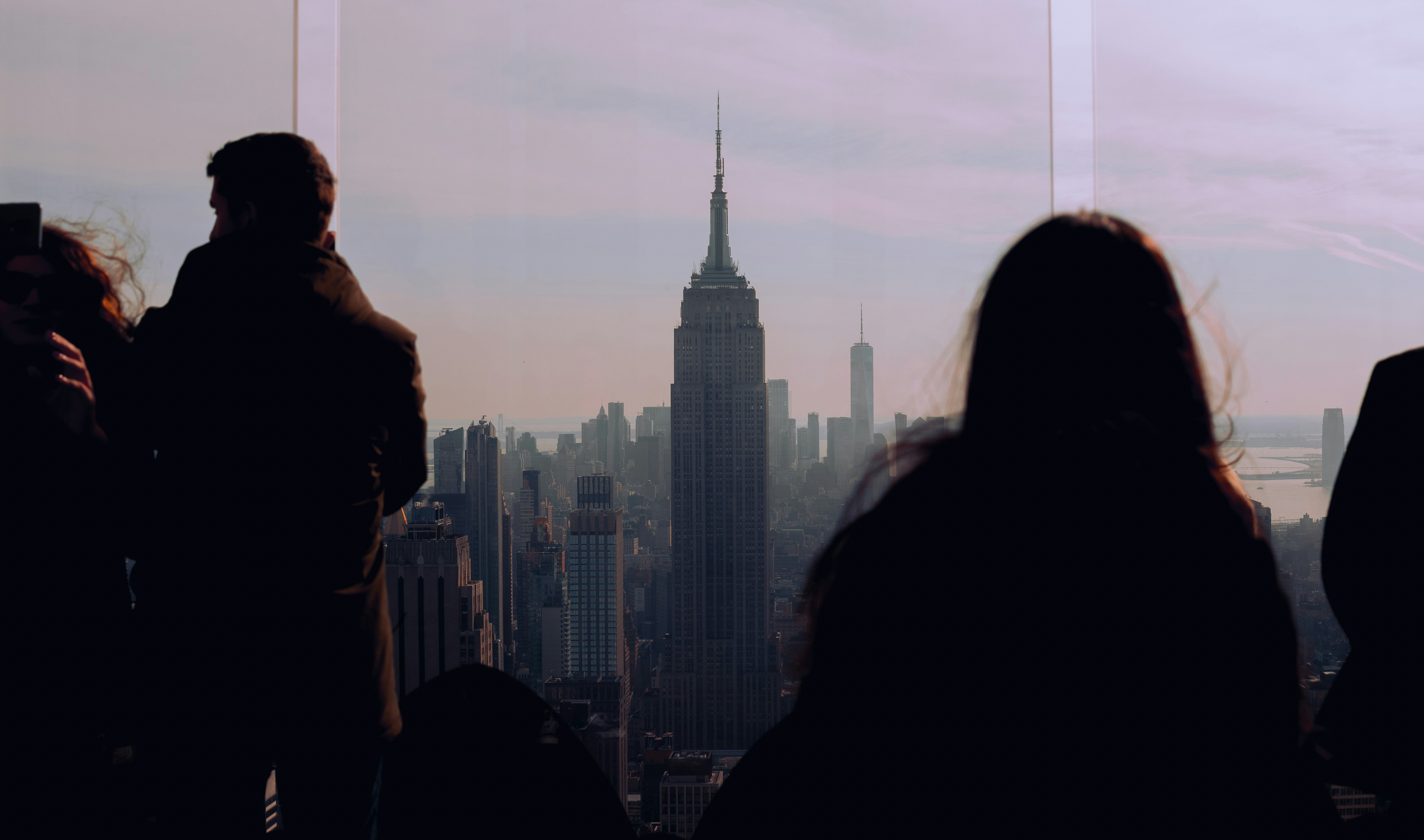 Silhouettes of visitors observing the New York City skyline with the Empire State Building prominently featured. A blend of urban exploration and human connection.