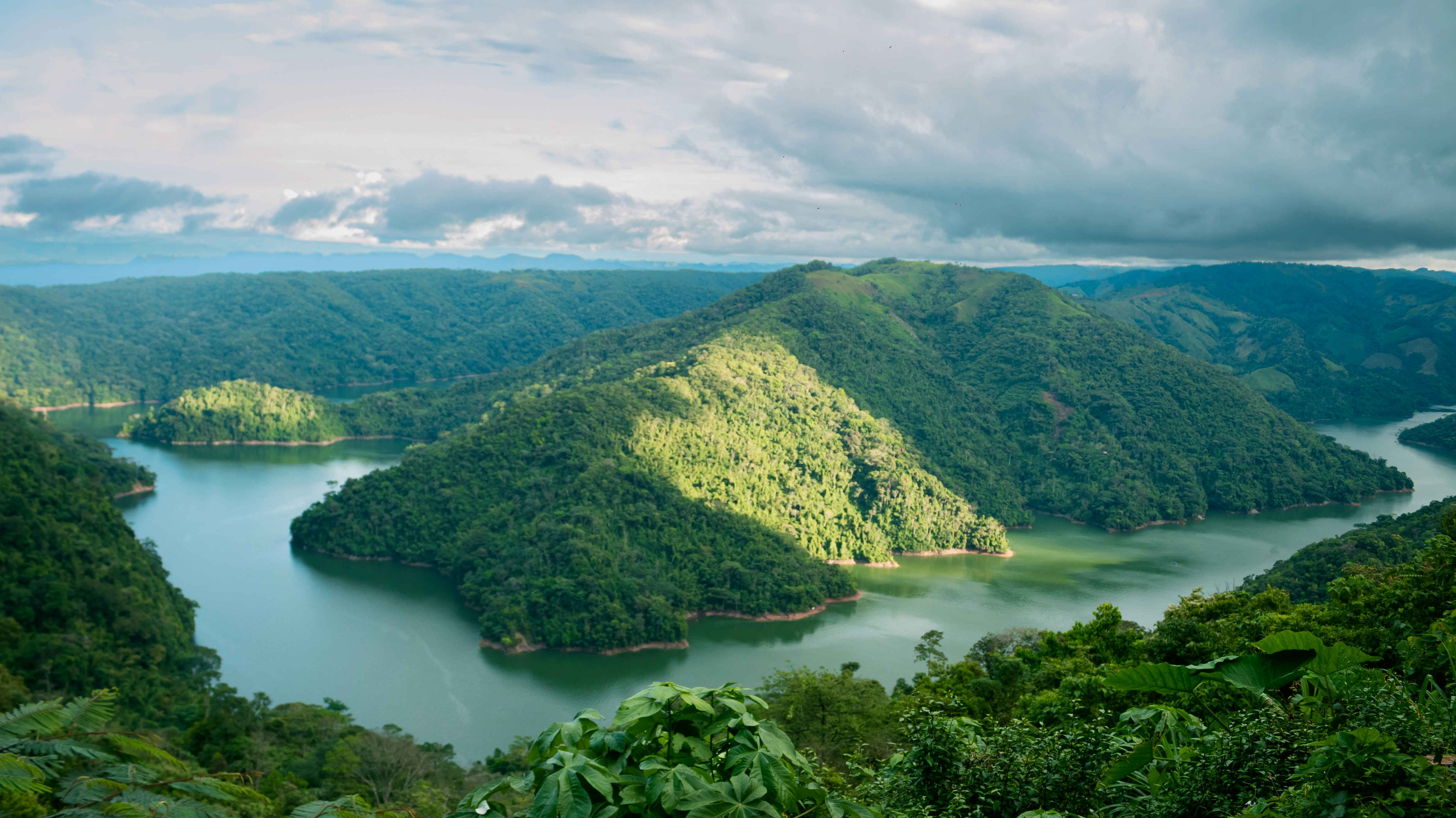 Lush green hills encircle a winding river under a cloud-filled sky.