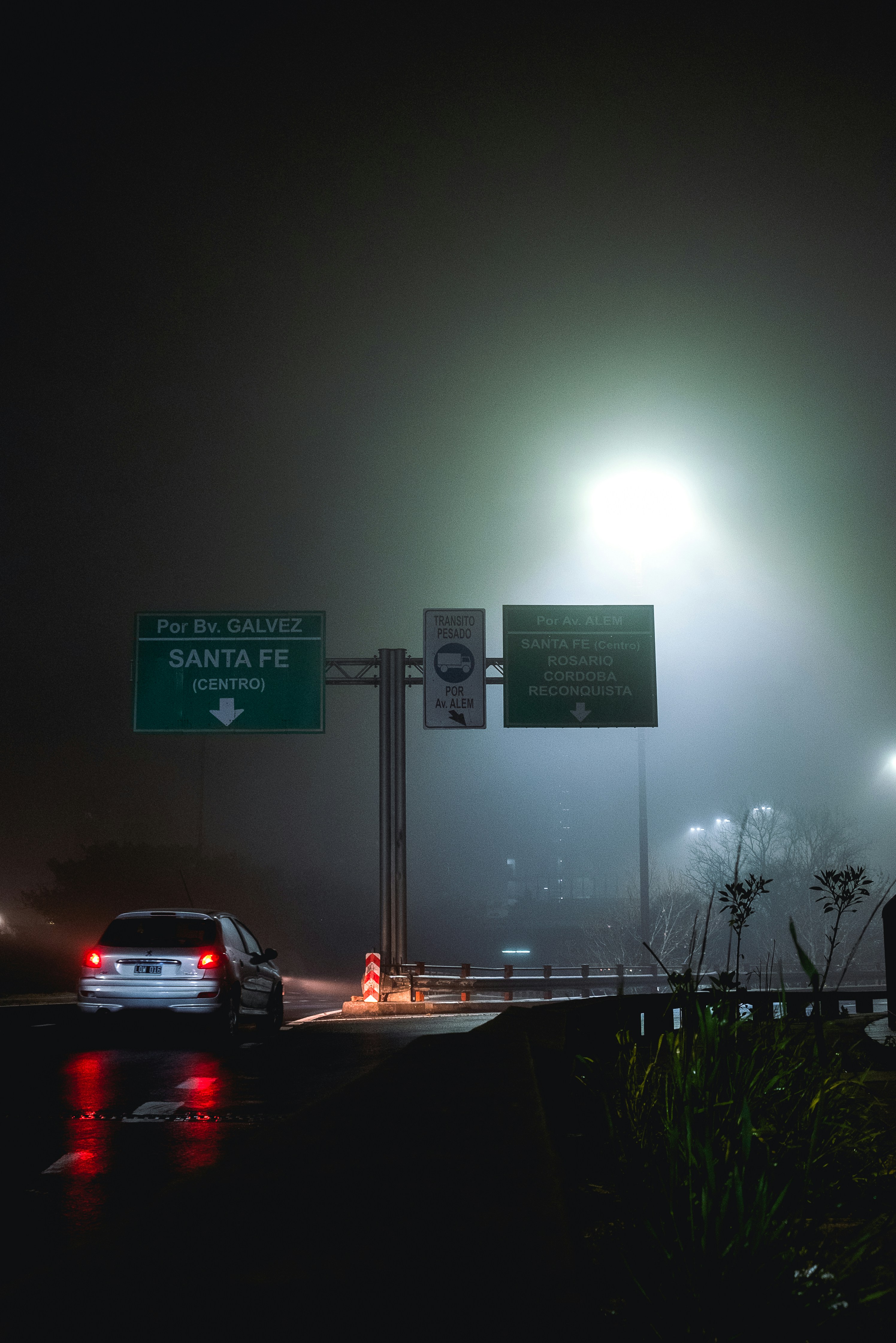 Dimly lit highway sign directing to Santa Fe, shrouded in thick fog with a car passing by. Wet pavement reflects the surrounding lights.