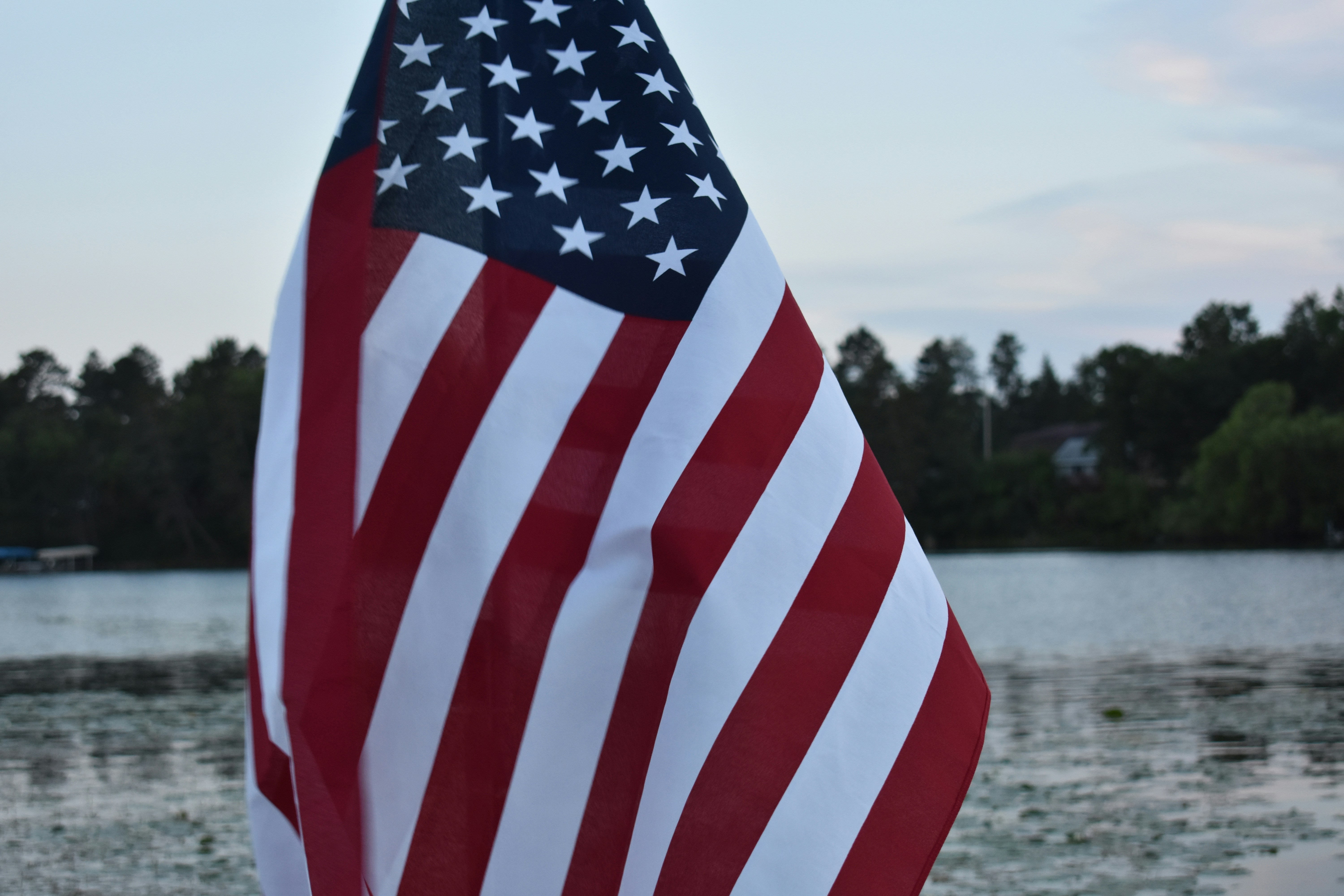 American flag fluttering gently by a serene lake, with trees lining the background. Captures a moment of tranquility and national pride.