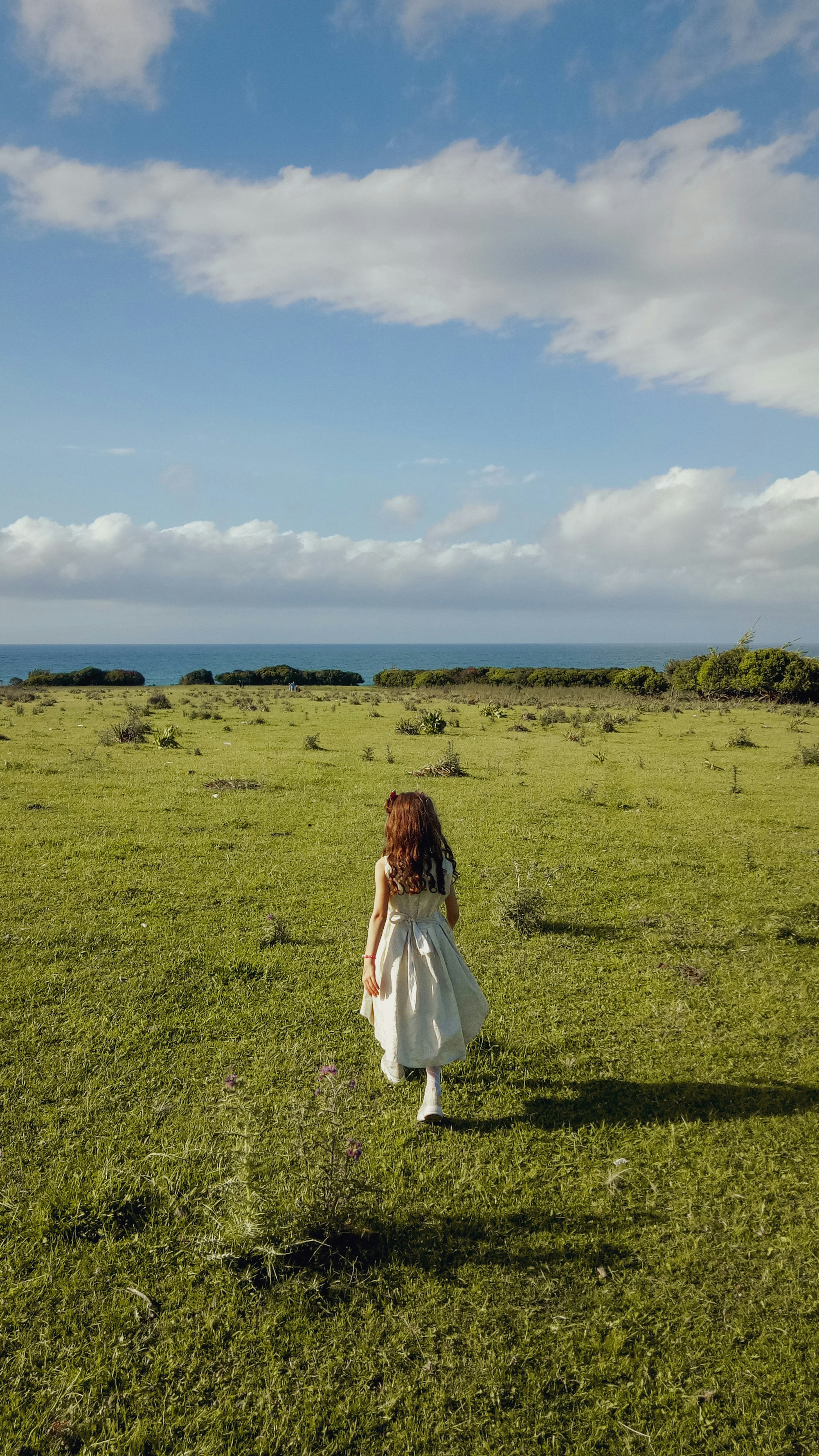A girl in a white dress walks away across a sunlit grassy field toward the distant sea under a blue sky.