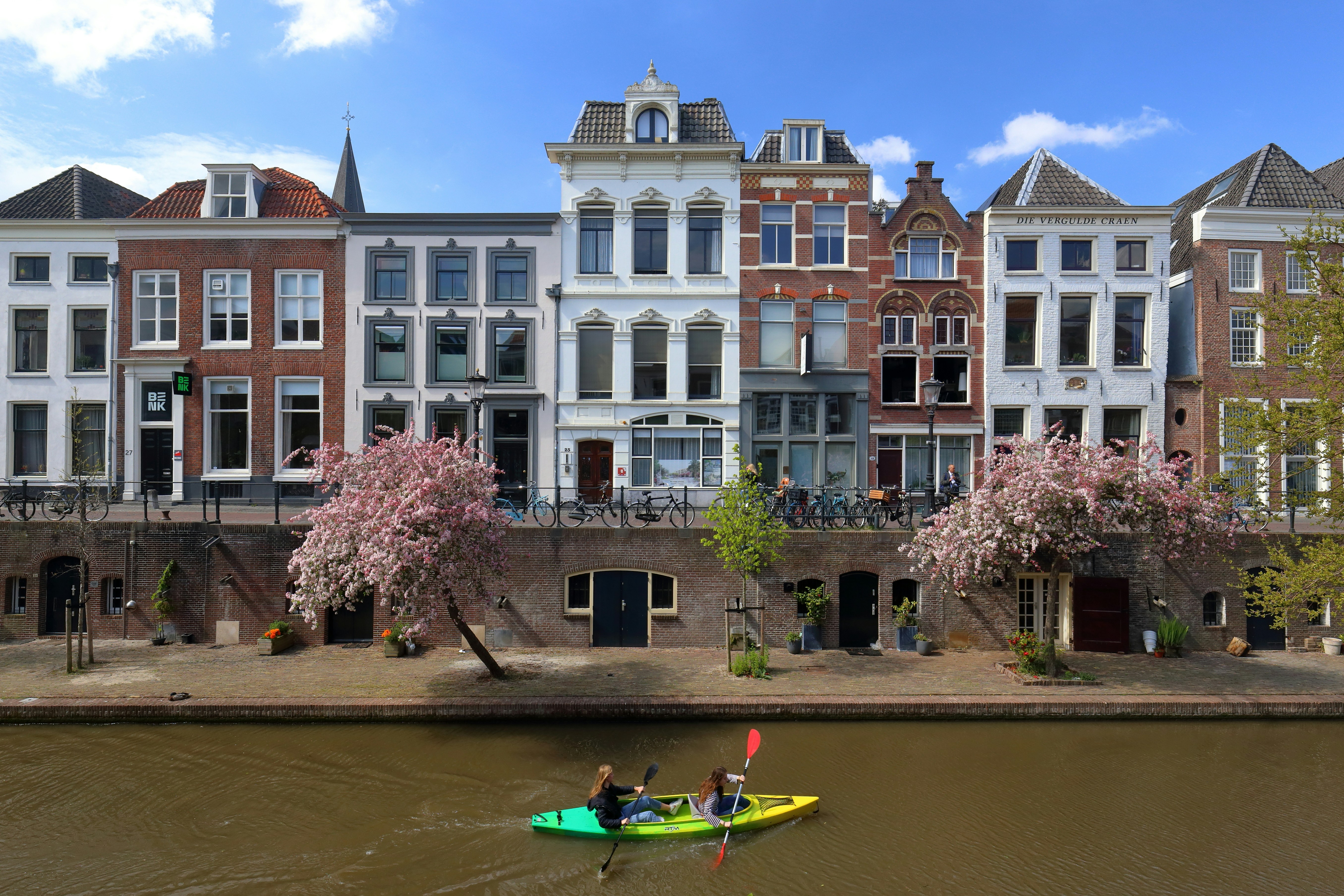 Two kayakers navigate a tranquil canal lined with charming historic buildings and blooming cherry trees.