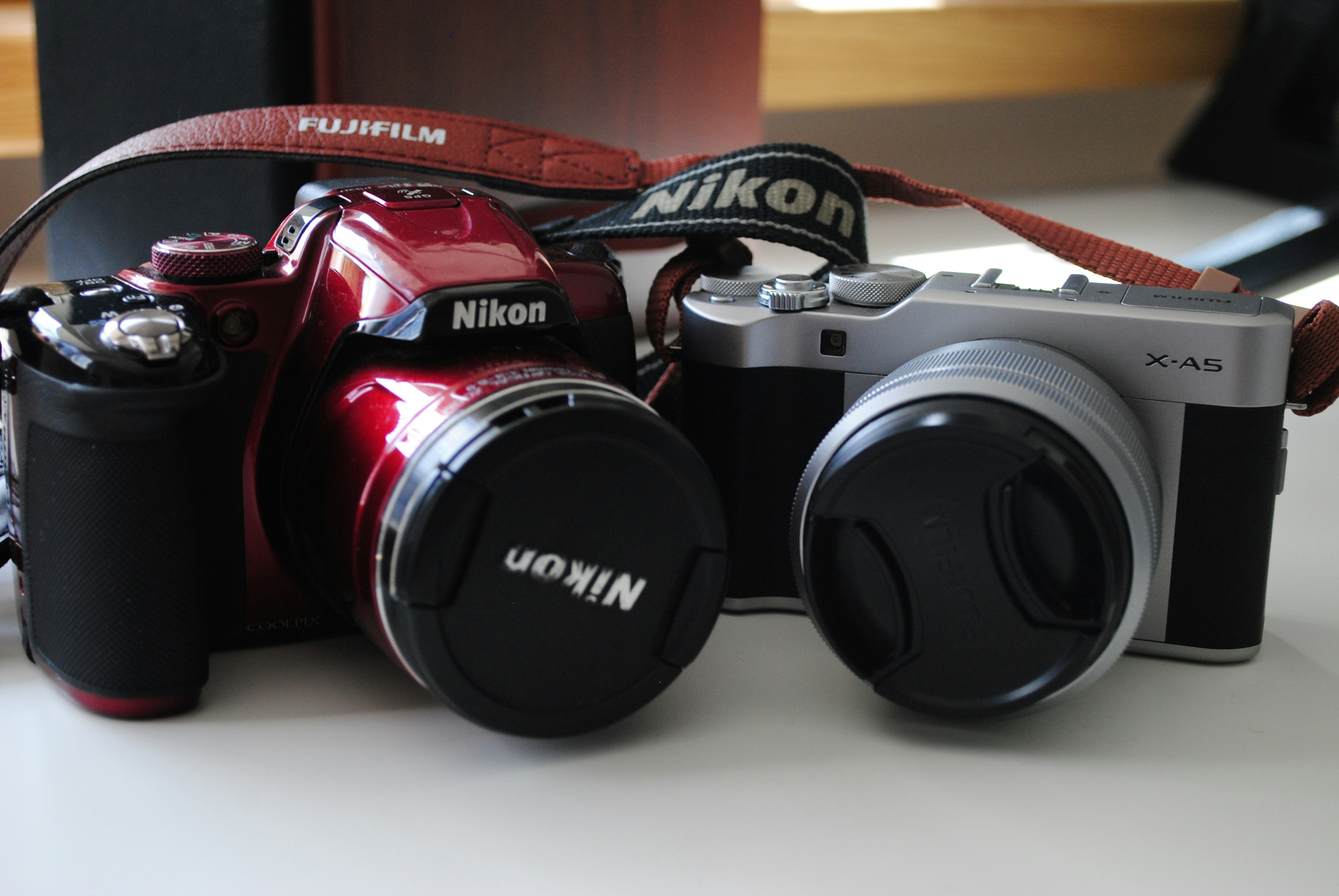 Japanese cameras on table at sunny day. Nikon P520 and Fujifilm X-A5.  | a red and black camera