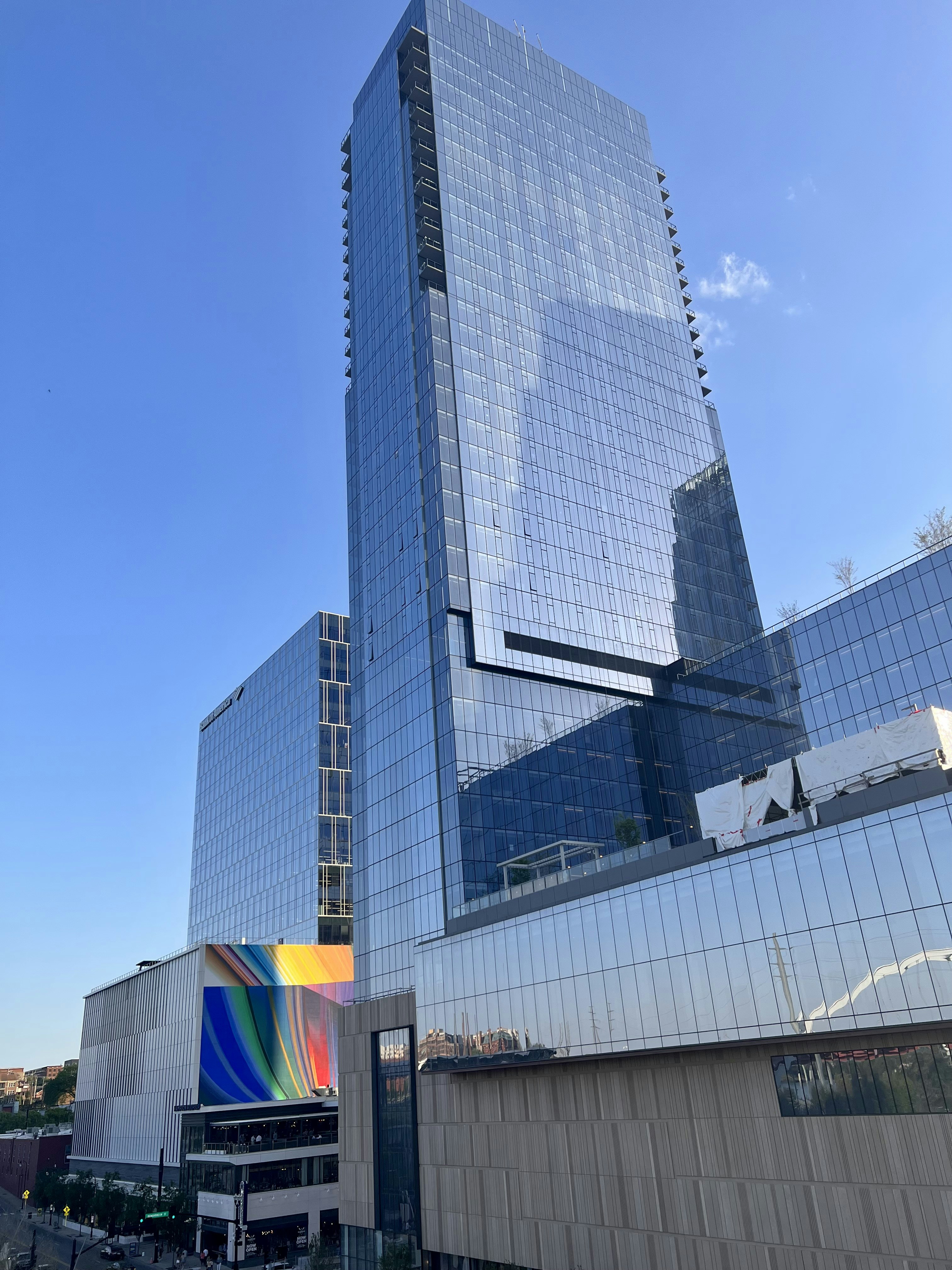 A sleek, glass-paneled skyscraper showcases vibrant artwork on its lower façade, reflecting the clear blue sky. The modern design emphasizes urban sophistication.