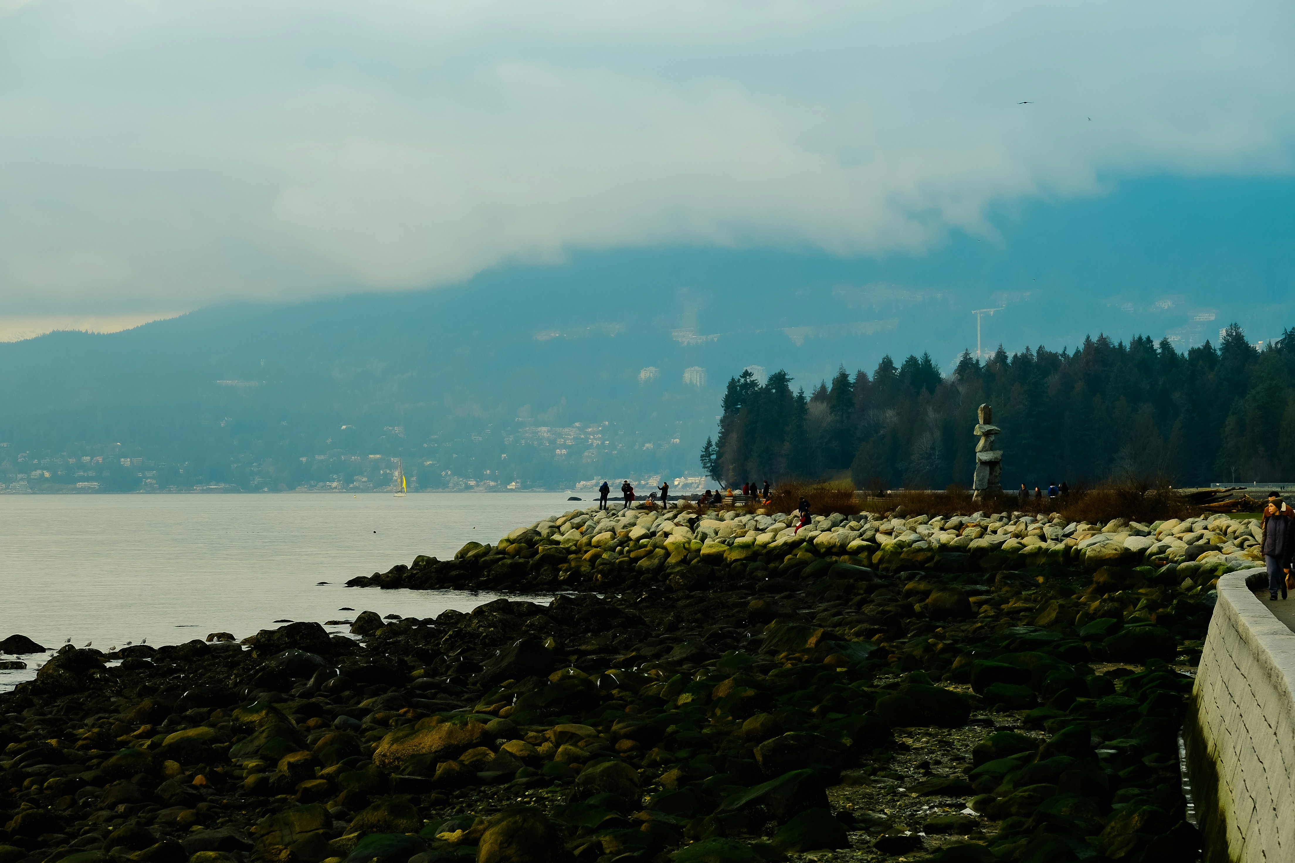 Rocky shoreline with a distant statue and people walking along the path, framed by a moody sky. The scene captures the essence of coastal leisure.