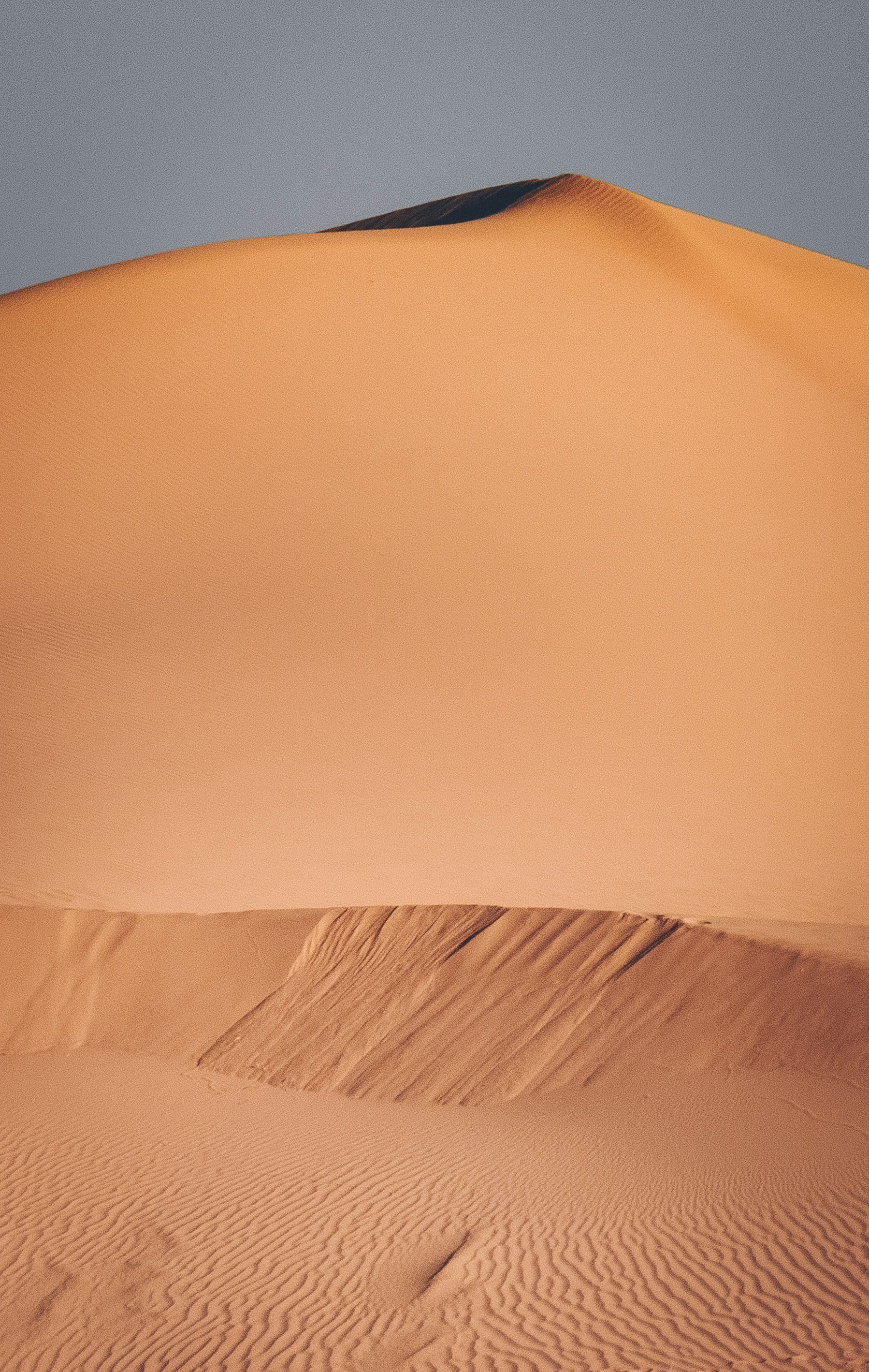 Gentle curves of a desert dune bathed in warm light, showcasing intricate patterns in the sand. The subtle gradient of color adds depth to the serene landscape.