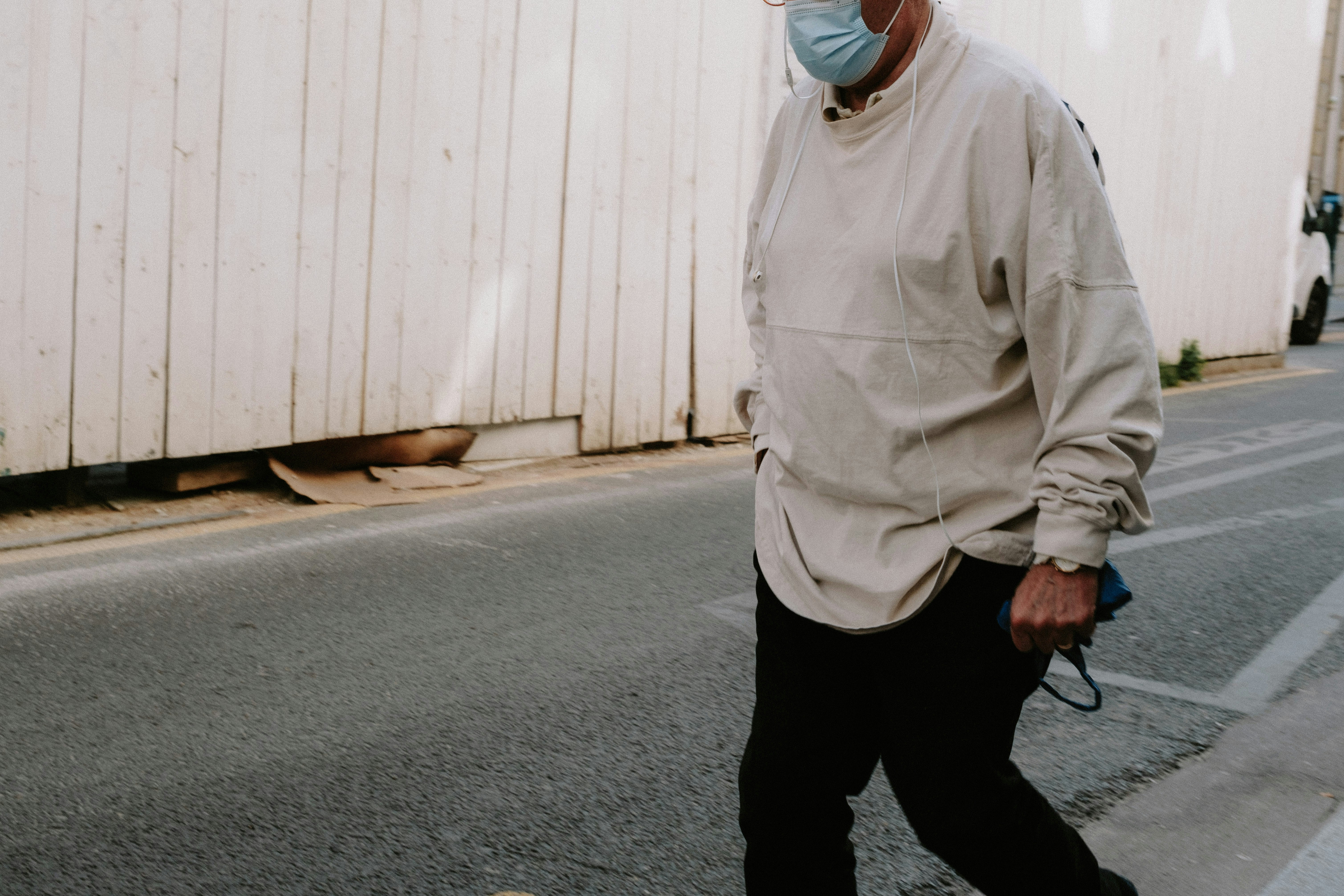 An individual in a light-colored sweater and mask walks purposefully along a city street, framed by a weathered wooden wall.