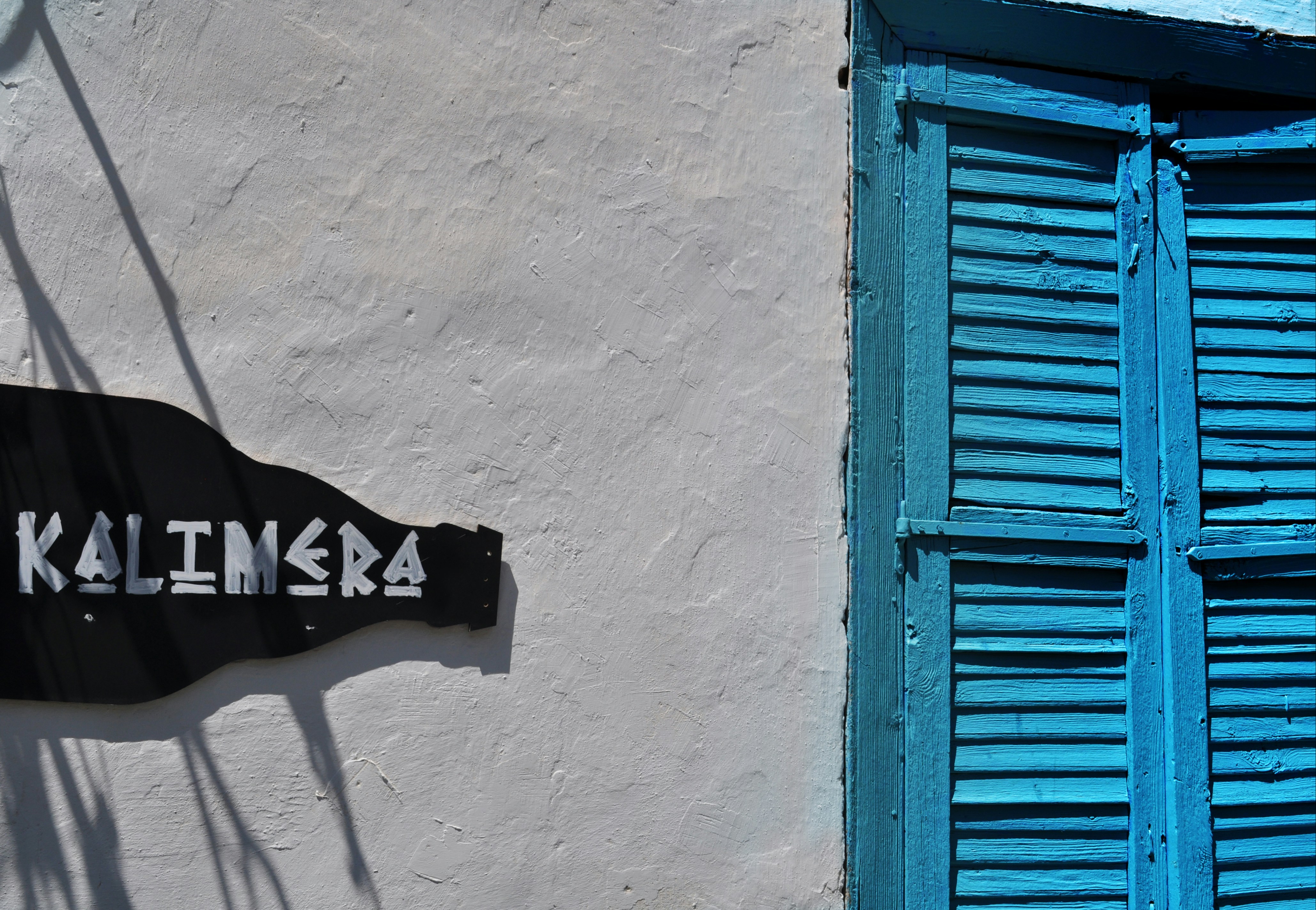KALIMERA on a Sunlit White Wall Framed by Electric Blue Shutters