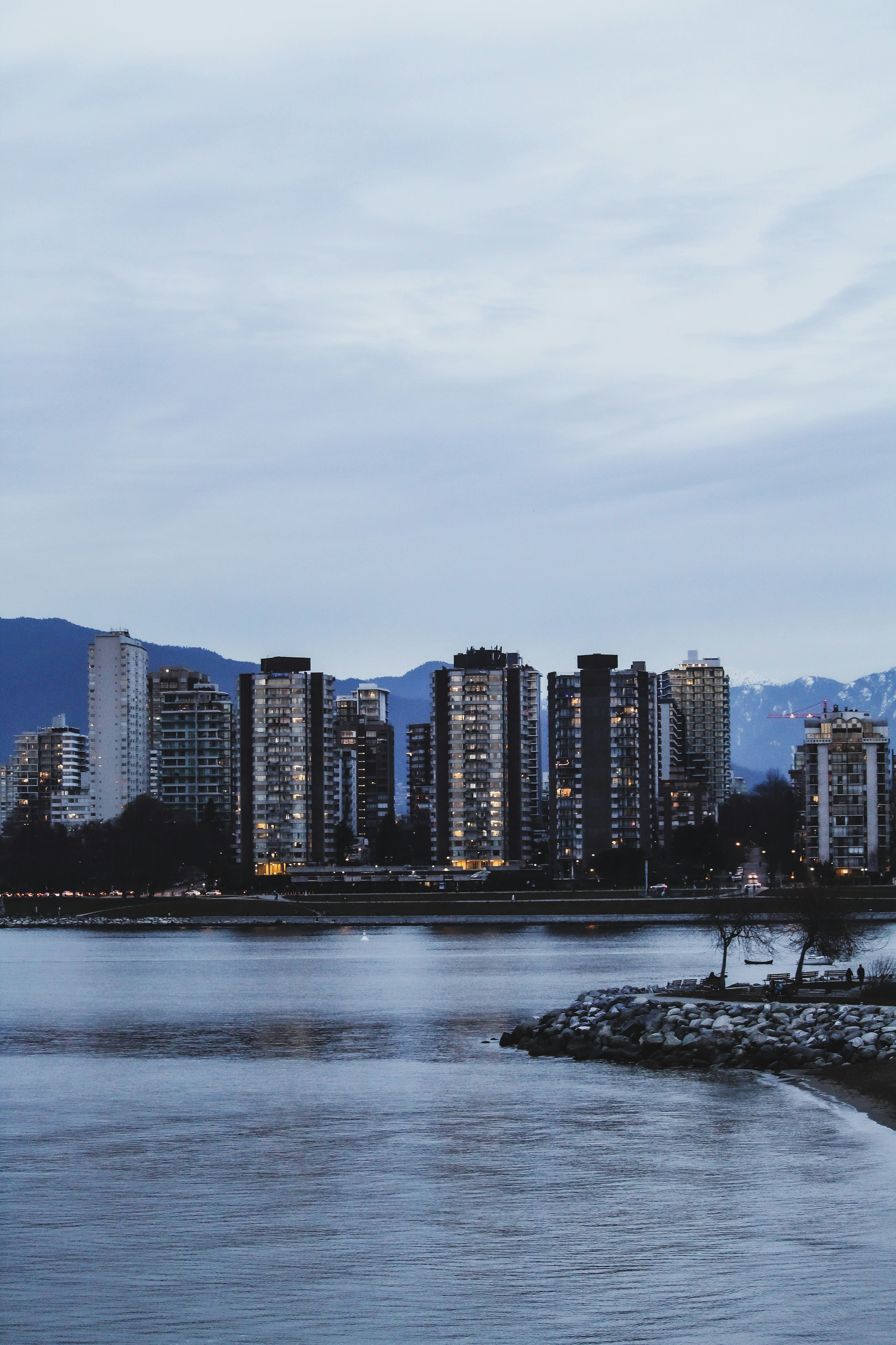 City skyline illuminated at dusk, reflecting on the calm waters below. The backdrop features distant mountains under a cloudy sky.