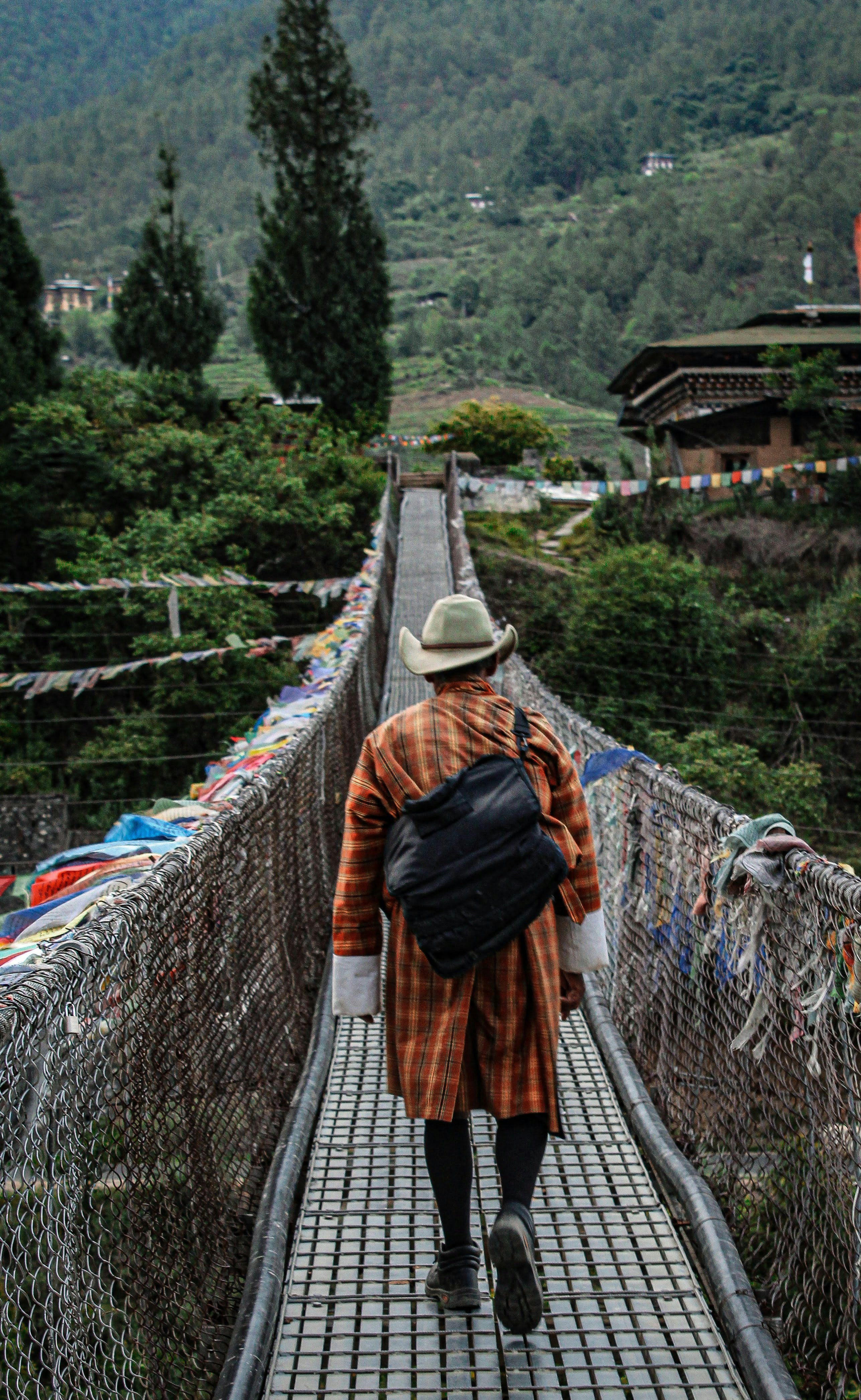 A traveler in traditional attire walks across a suspension bridge adorned with colorful prayer flags, surrounded by lush greenery and distant mountains.