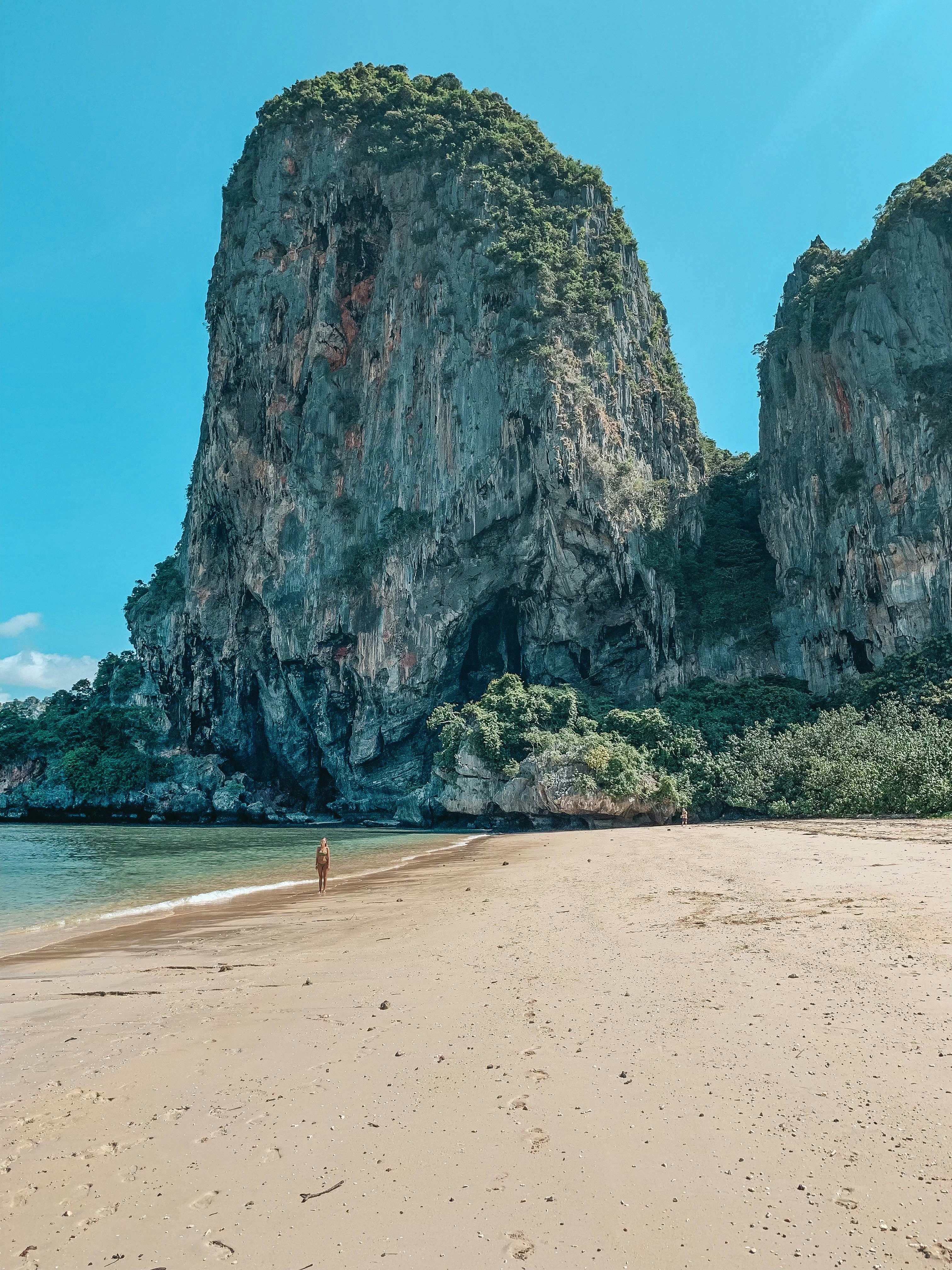 Towering limestone cliffs rise dramatically above a serene beach, with a solitary figure walking along the shore. The clear blue sky enhances the tranquil atmosphere.