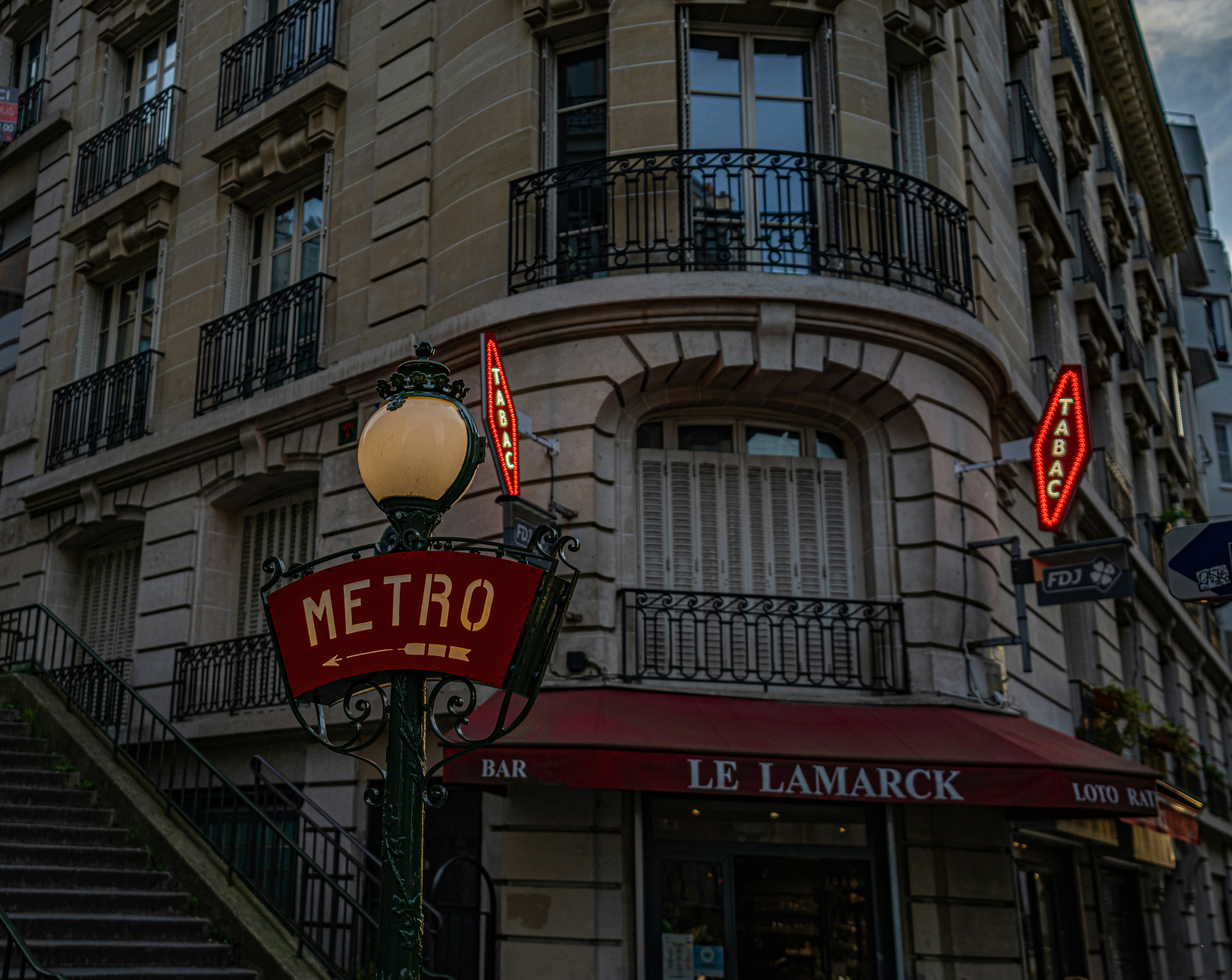 Vintage Metro sign stands prominently in front of a classic Parisian building with illuminated storefronts. The scene captures the essence of urban life in Paris.