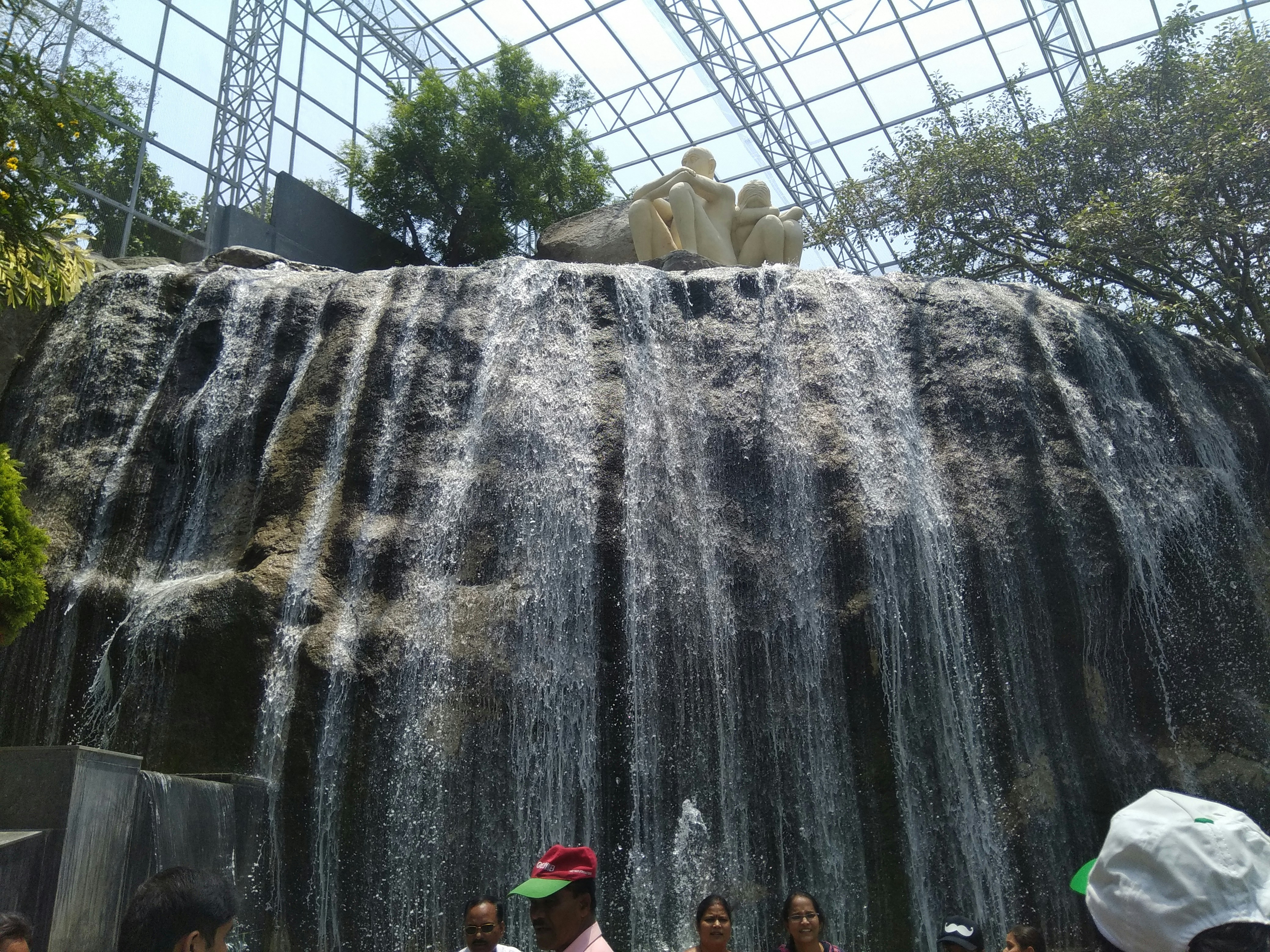 A serene waterfall cascades down a rocky surface, framed by lush greenery and a glass canopy. Two sculpted figures sit peacefully atop the waterfall, blending art and nature.