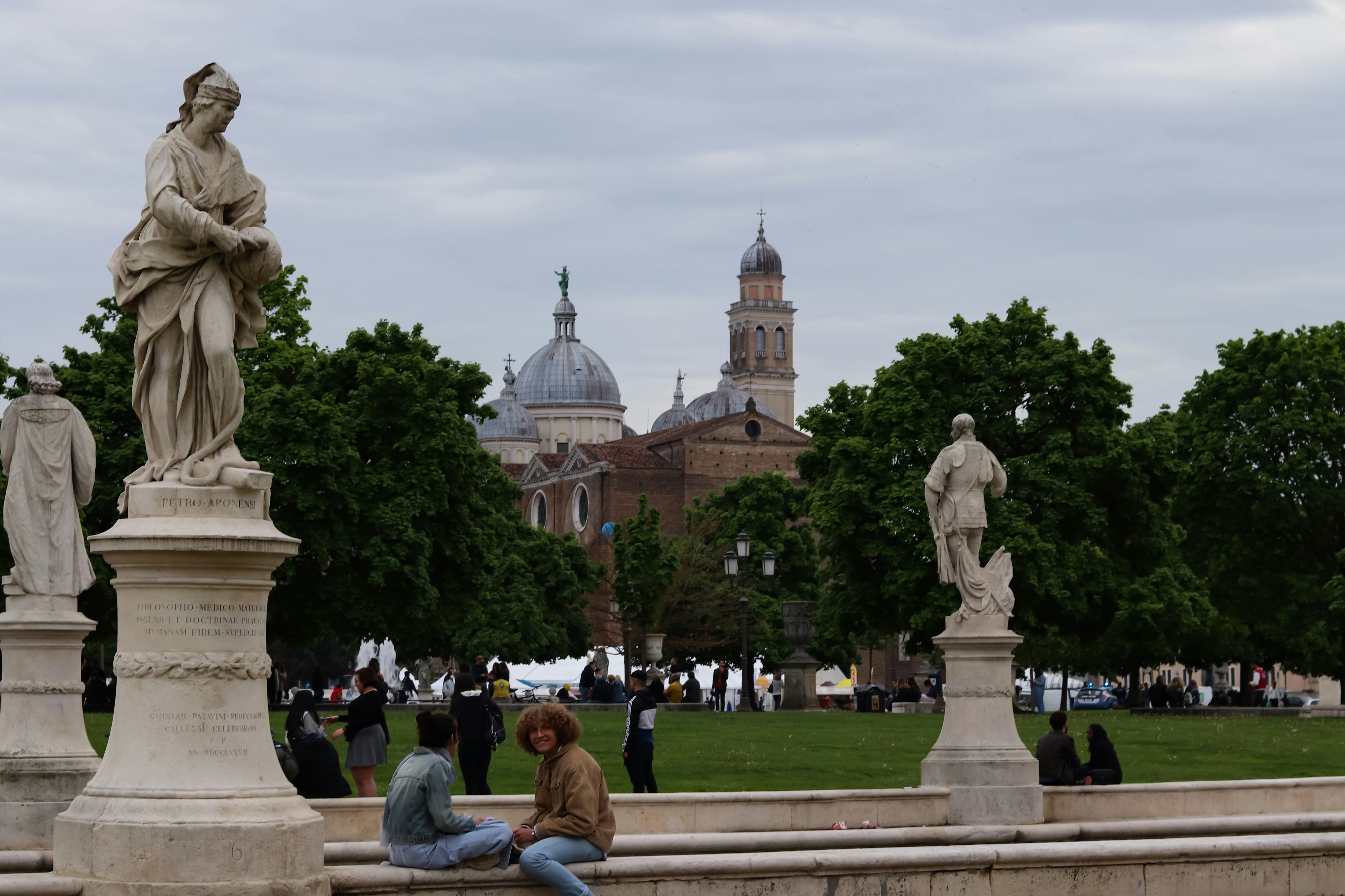 a group of people sitting on a grass field with statues in front of a building